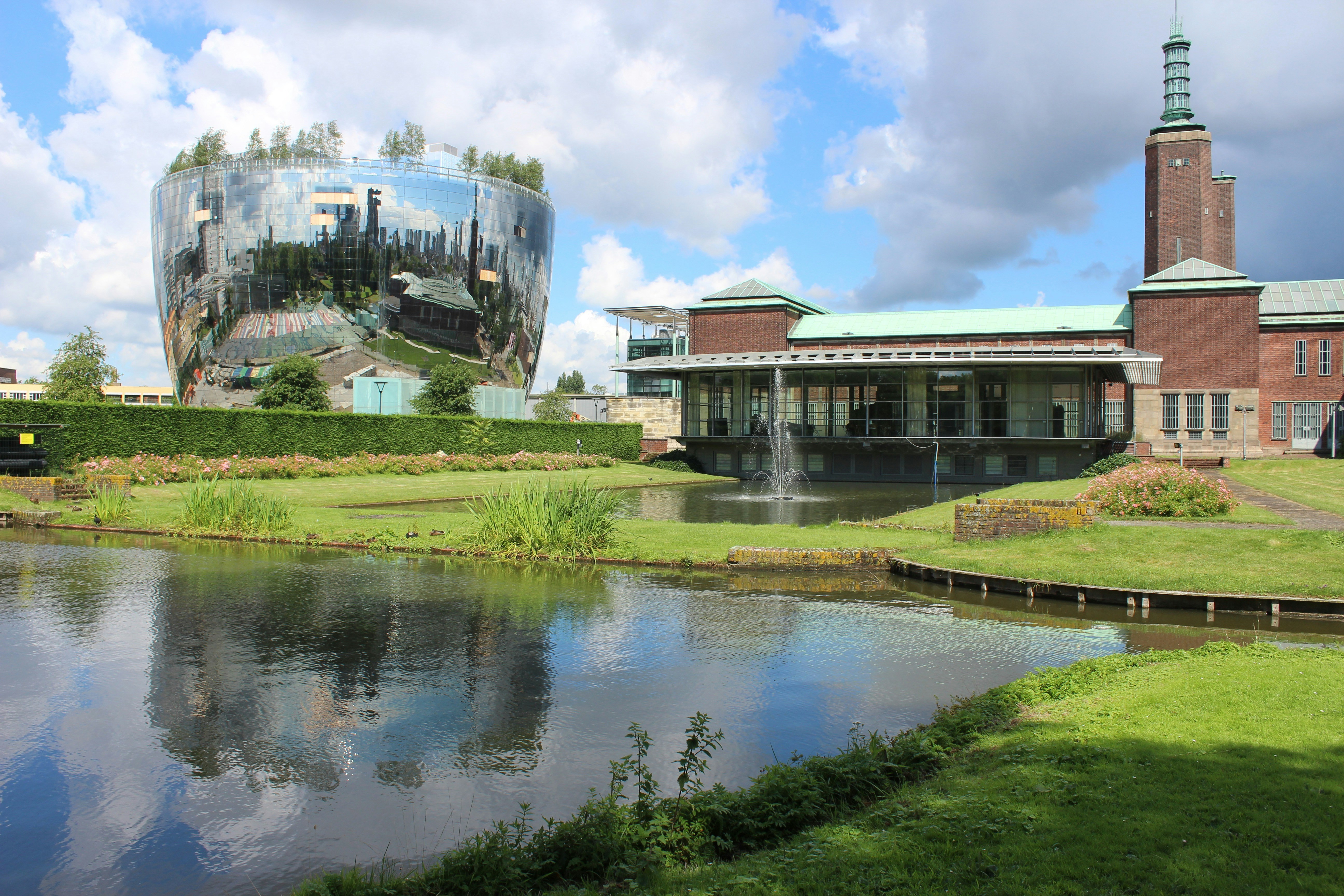 Modern mirrored building and historic museum near a serene pond under a partly cloudy sky.