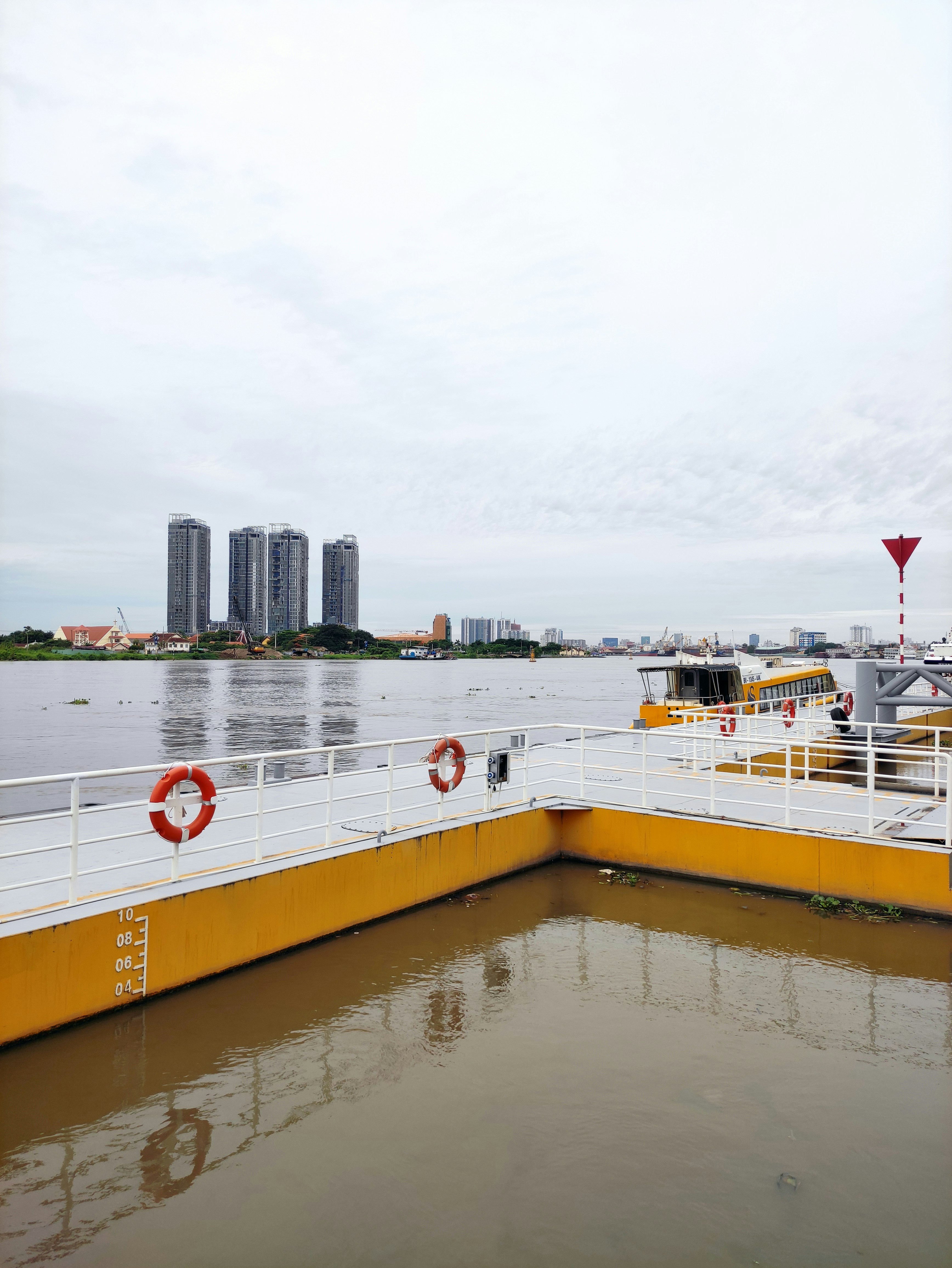 Photo of a riverside pier with bold yellow railing framing calm water and a distant city skyline, with a small yellow boat on the right.