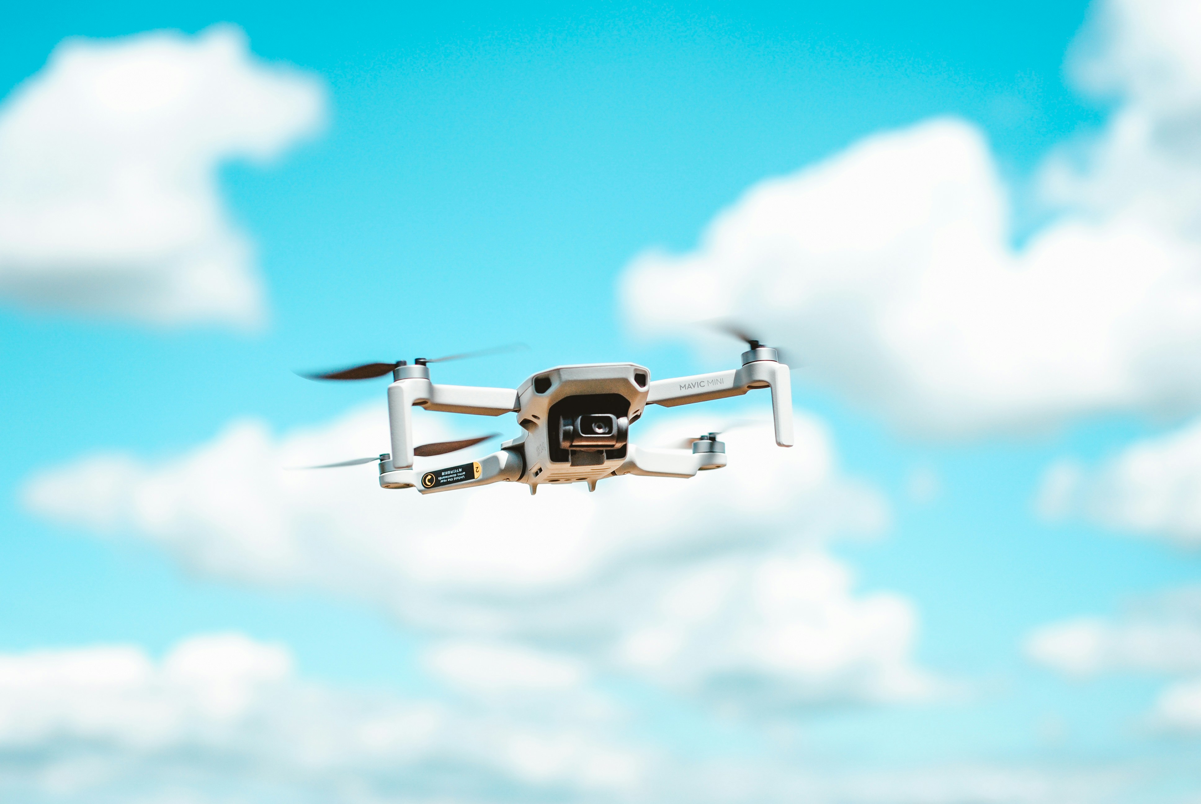 A drone soaring against a backdrop of fluffy white clouds in a clear blue sky.