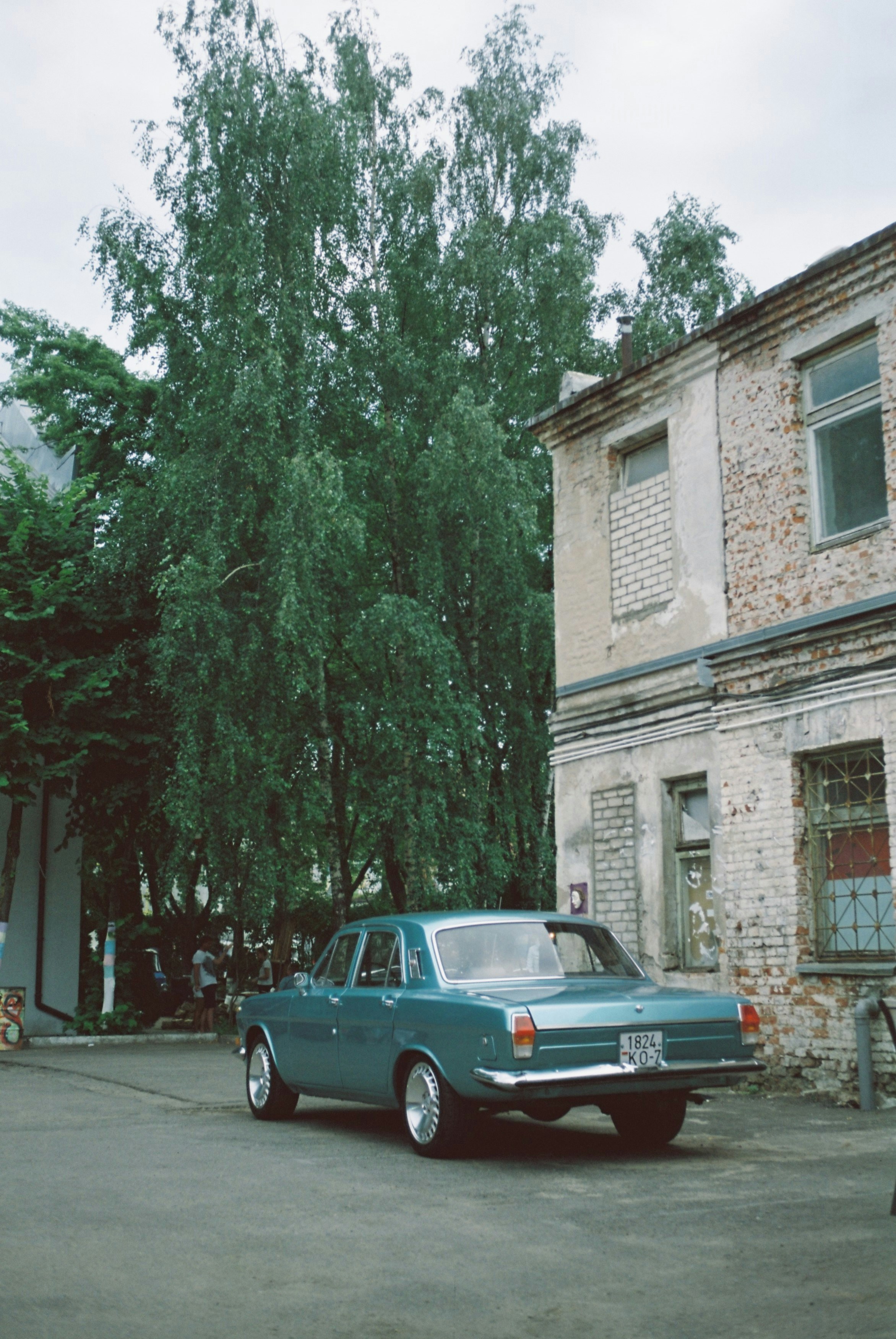 Vintage car parked beside an old brick building, surrounded by lush trees in an urban setting.