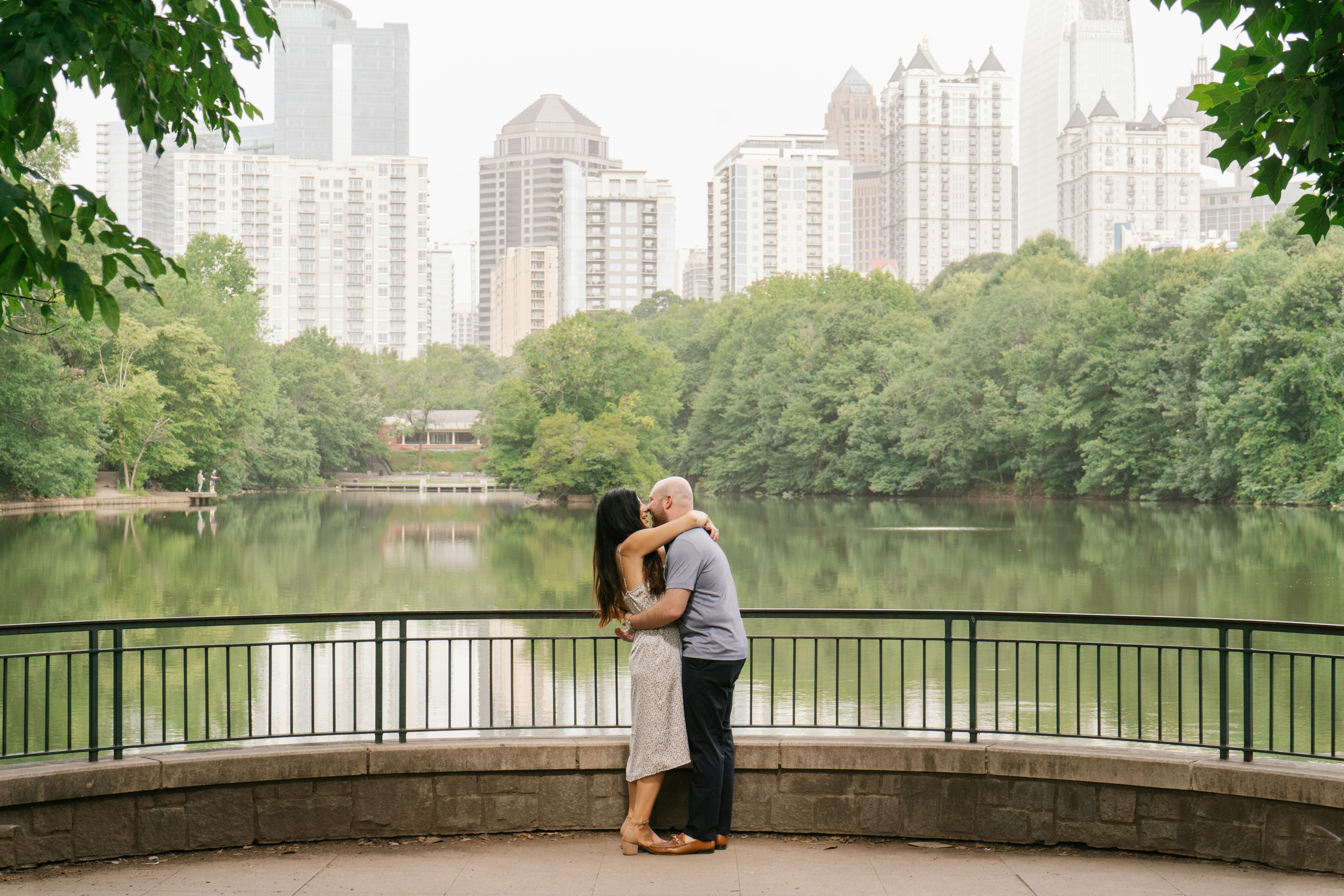 Couple embracing near a tranquil lake with a city skyline in the background, framed by lush green trees.