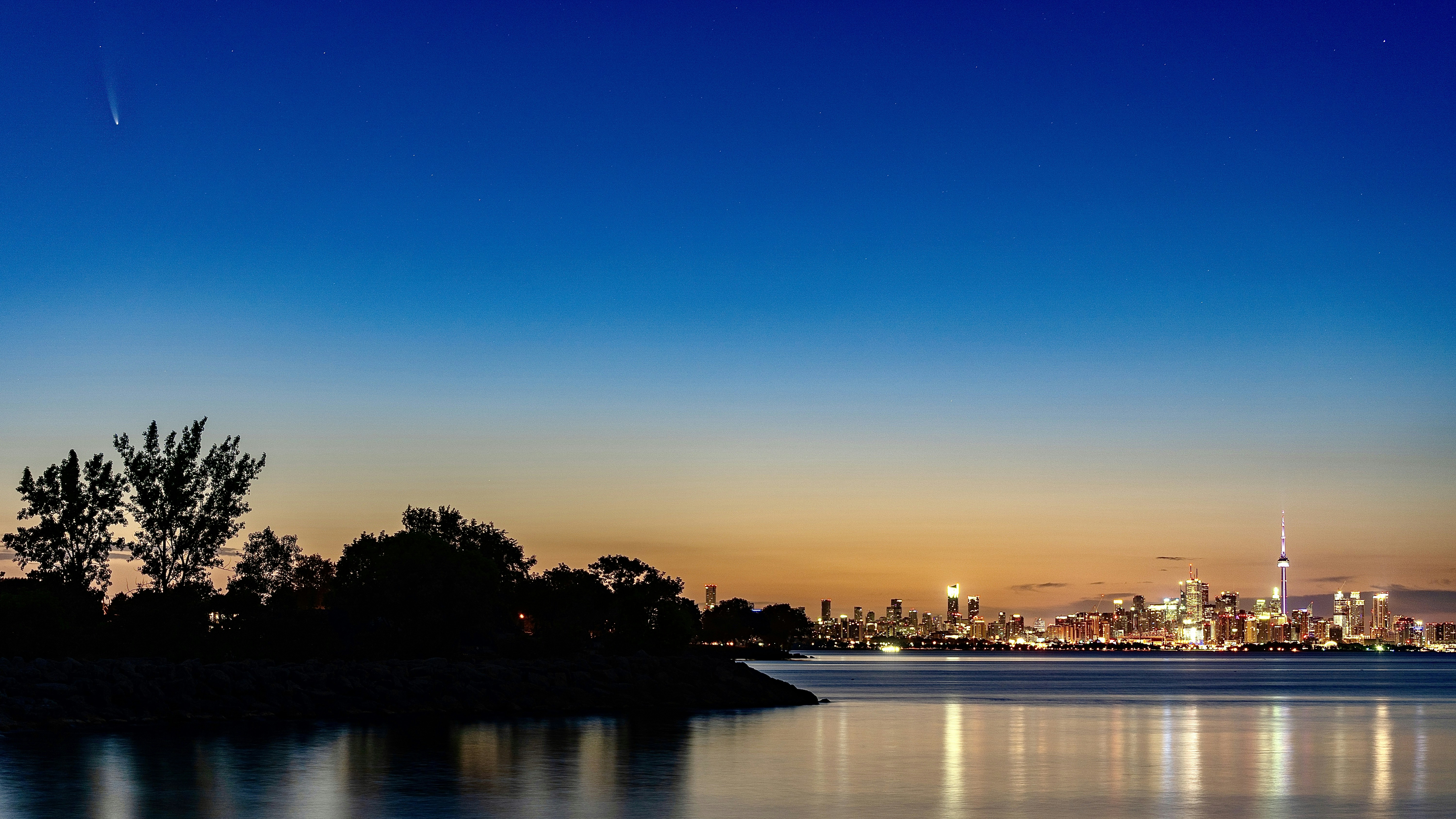 A comet streaks across the twilight sky above a tranquil waterfront, with a city skyline illuminated in the distance. The scene captures the serene transition from day to night.
