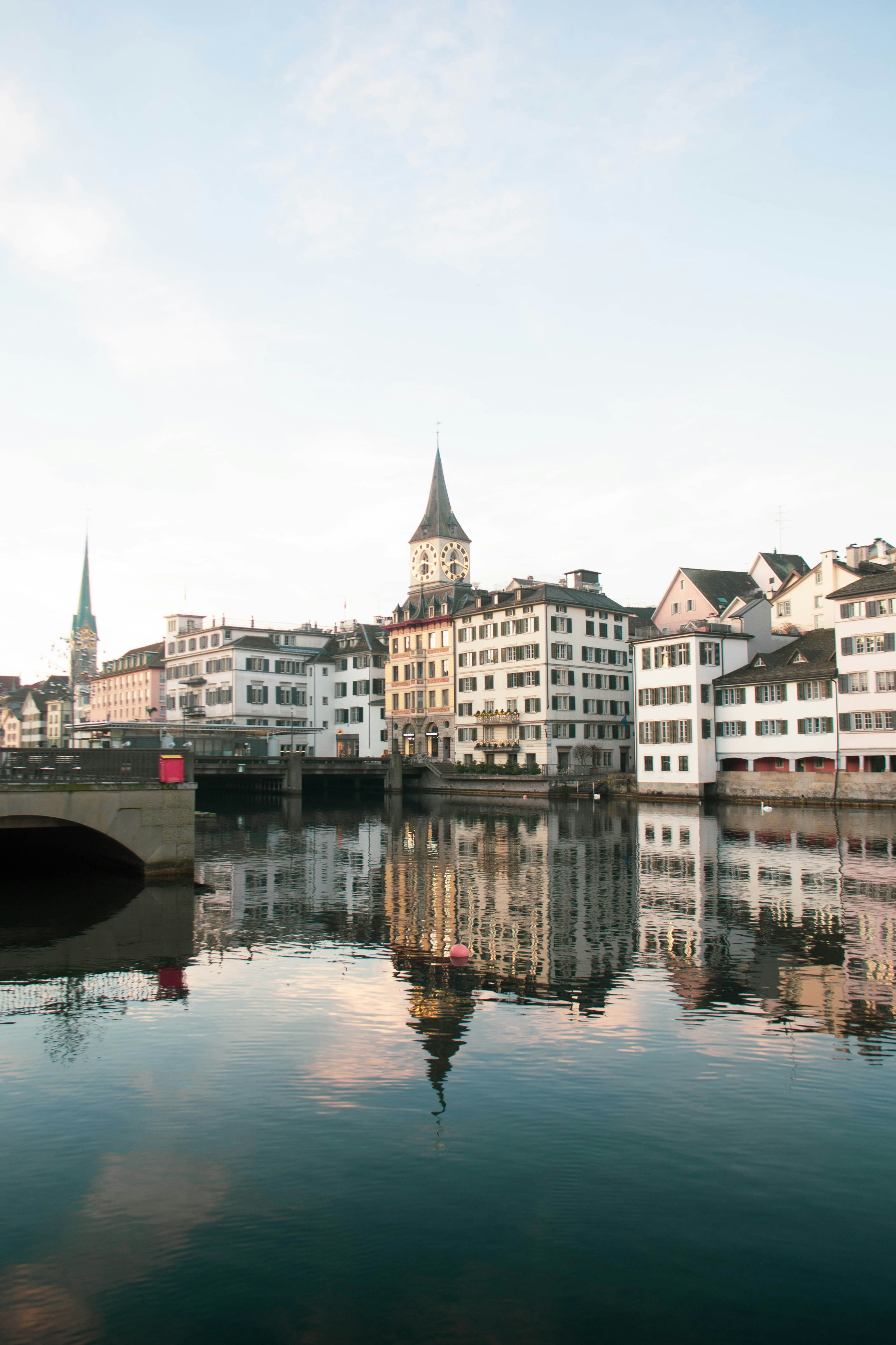 Historic buildings lining the riverbank of Zurich, with their reflections mirrored on the calm water. A tranquil scene showcasing the city's architectural charm.