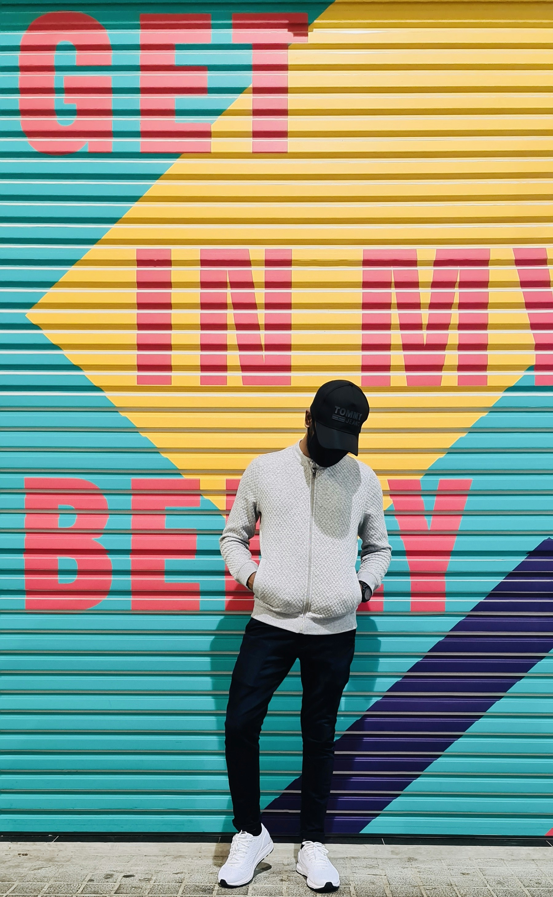 A man wearing a light gray hoodie, dark pants, white sneakers, and a black cap stands with hands in pockets in front of a bold geometric mural painted on a metal shutter.