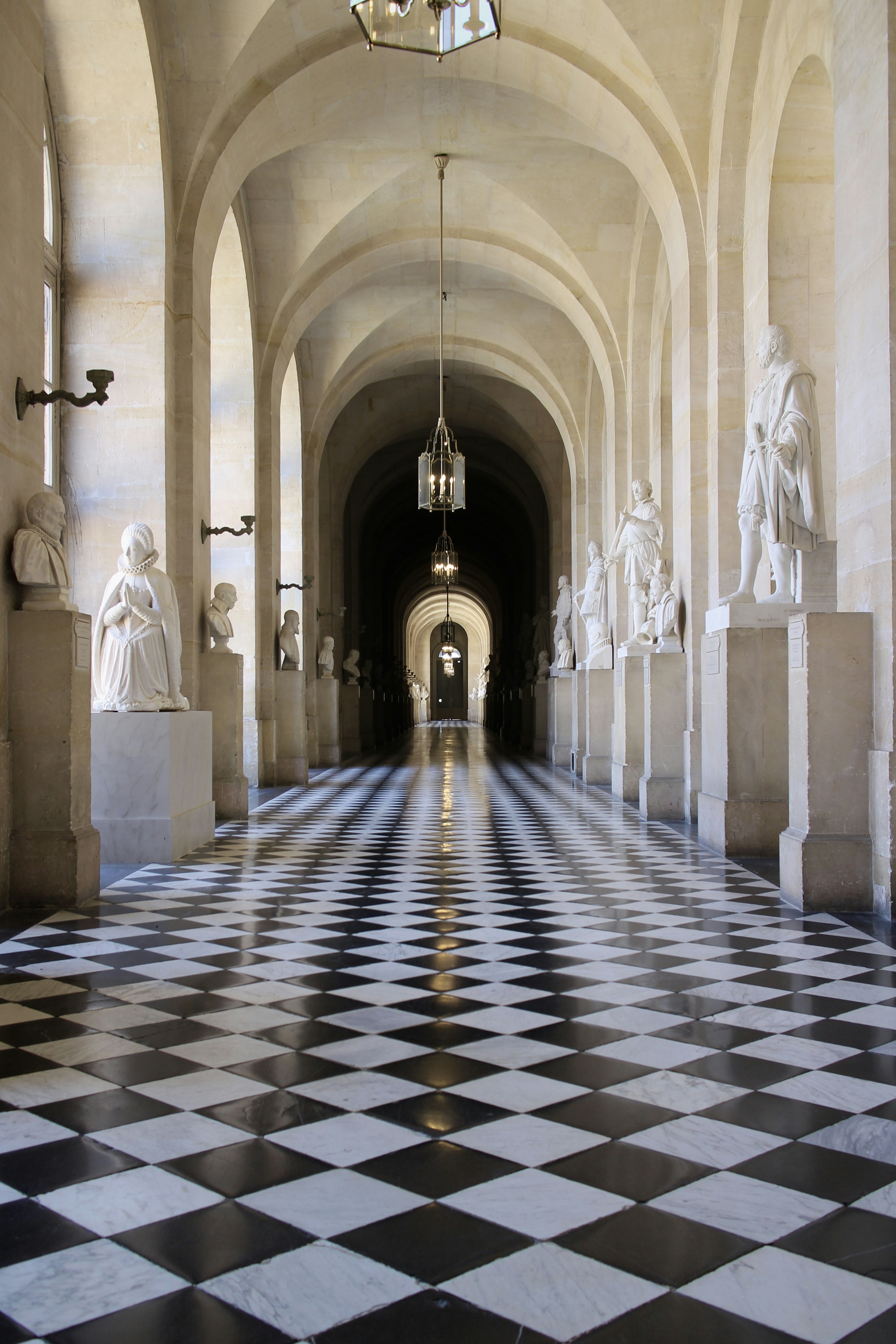 A grand corridor lined with marble statues, featuring a striking black and white checkered floor and elegant arches. The soft lighting enhances the architectural details.