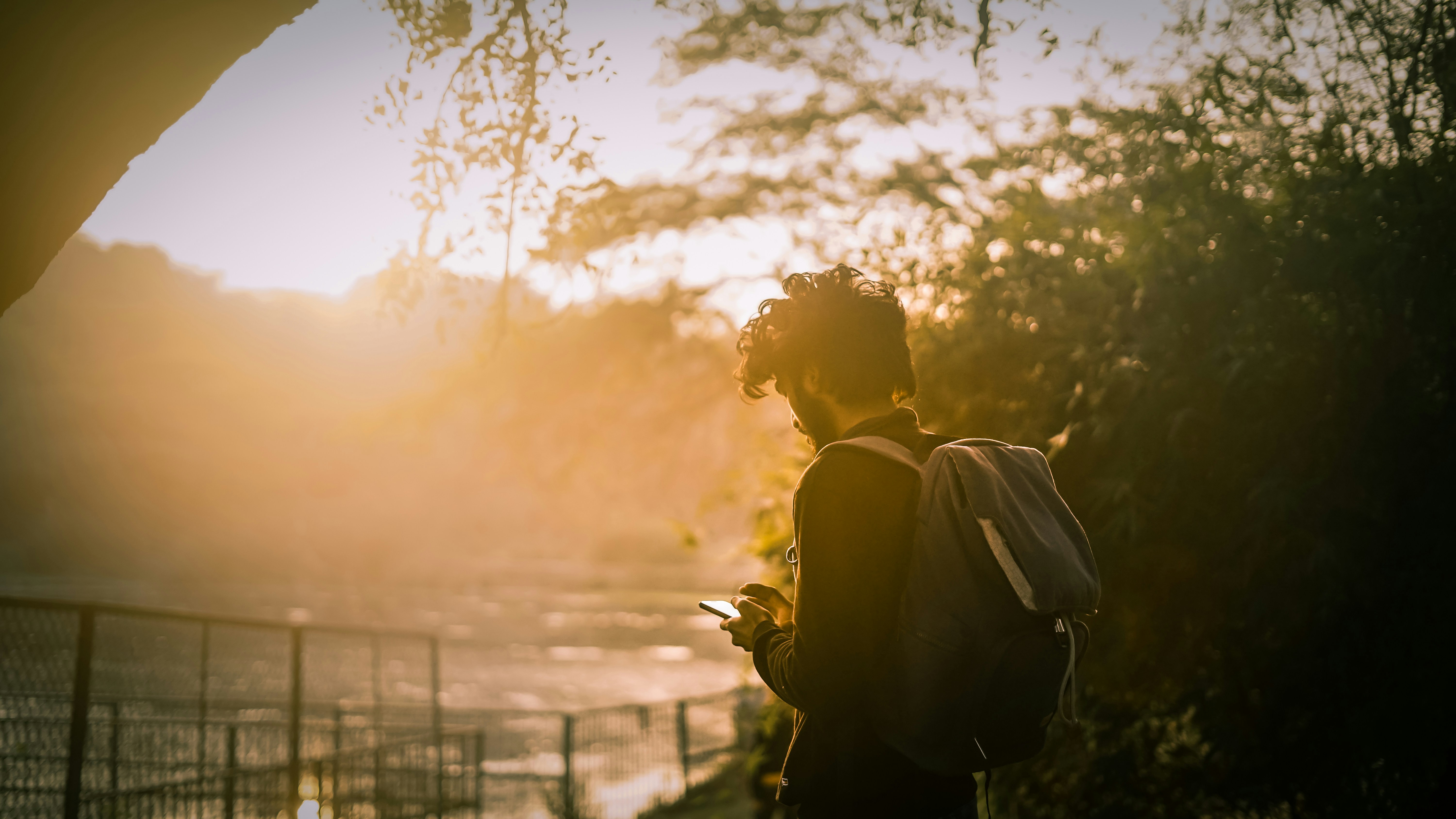 Golden Hour Silhouette: Backpacked Traveler Checks a Phone by a Sunlit Riverside Rail