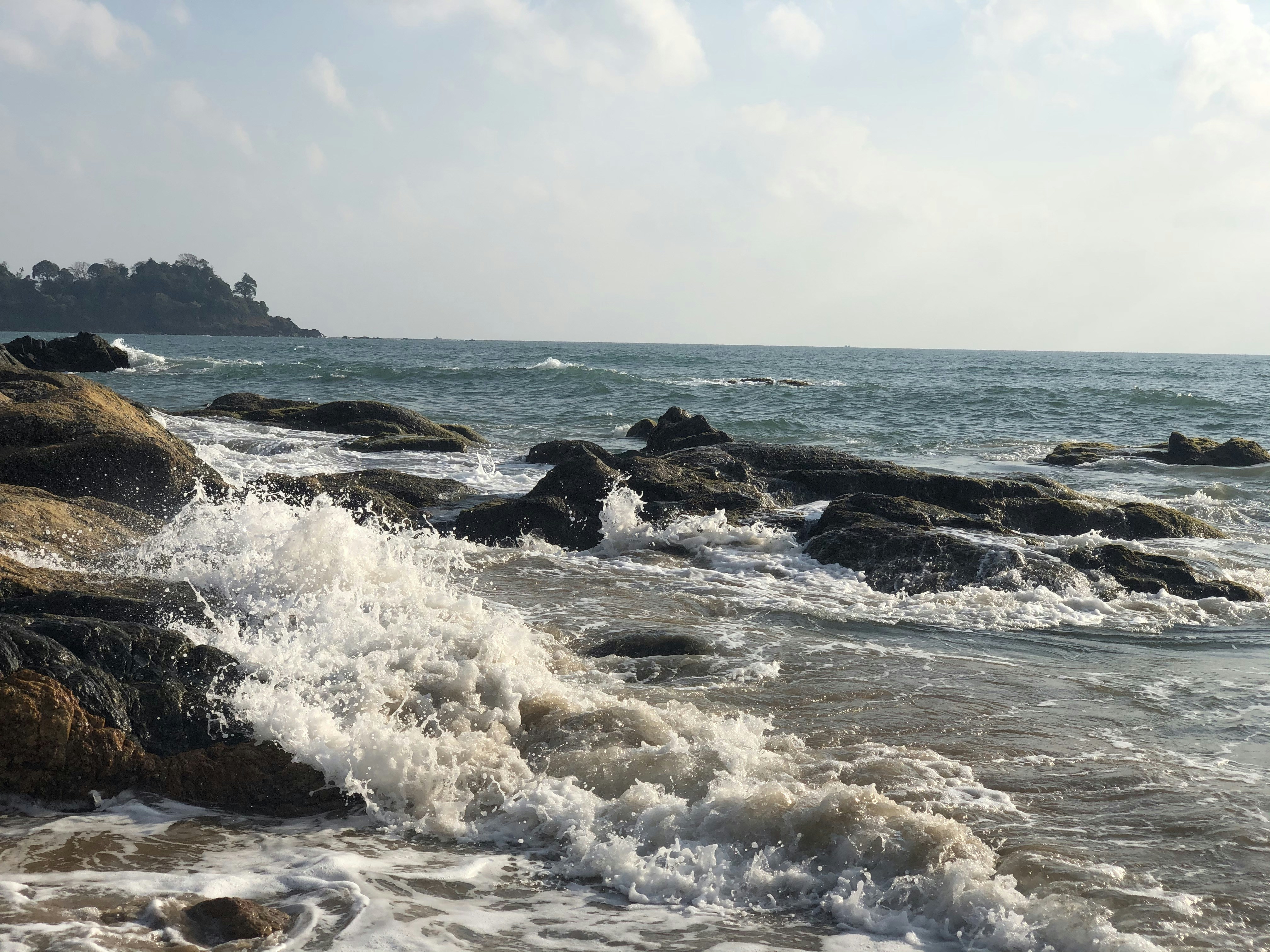 Foamy waves crashing against rugged rocks along a serene coastline under a cloudy sky.