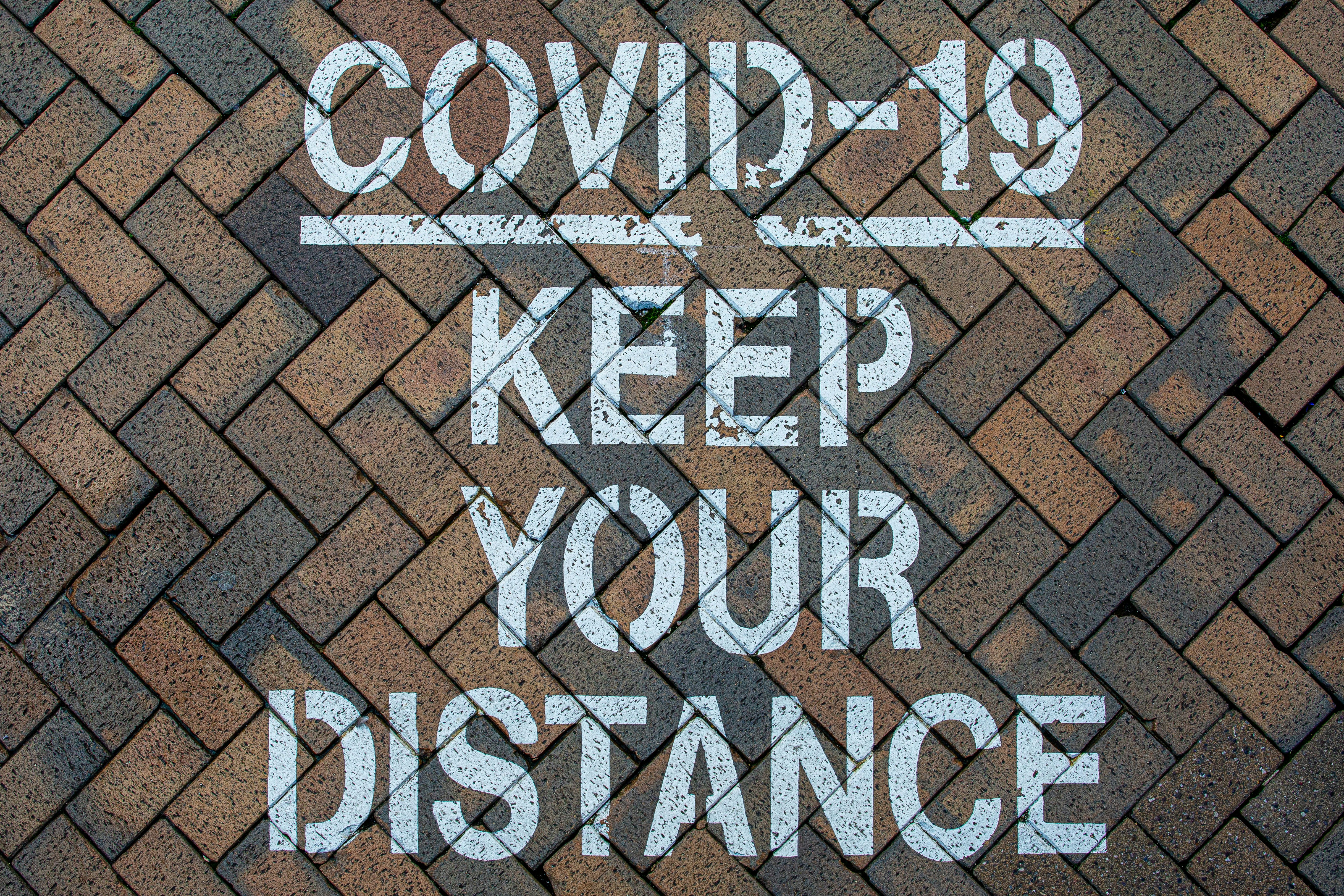 Stencil-style text on brick pavement urging social distancing during COVID-19.