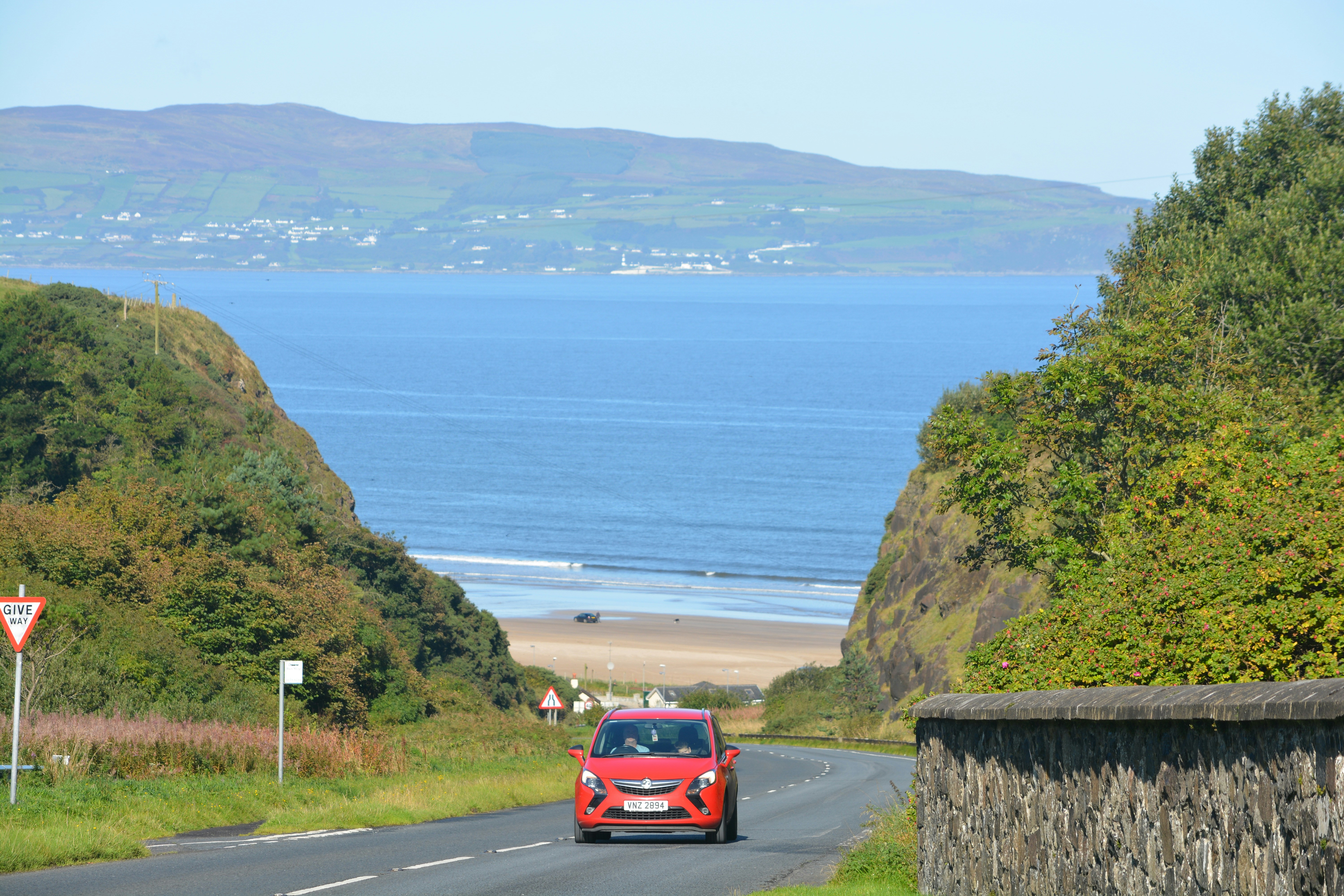 Red Hatchback Traverses a Cliffside Coastal Road Toward a Secluded Beach