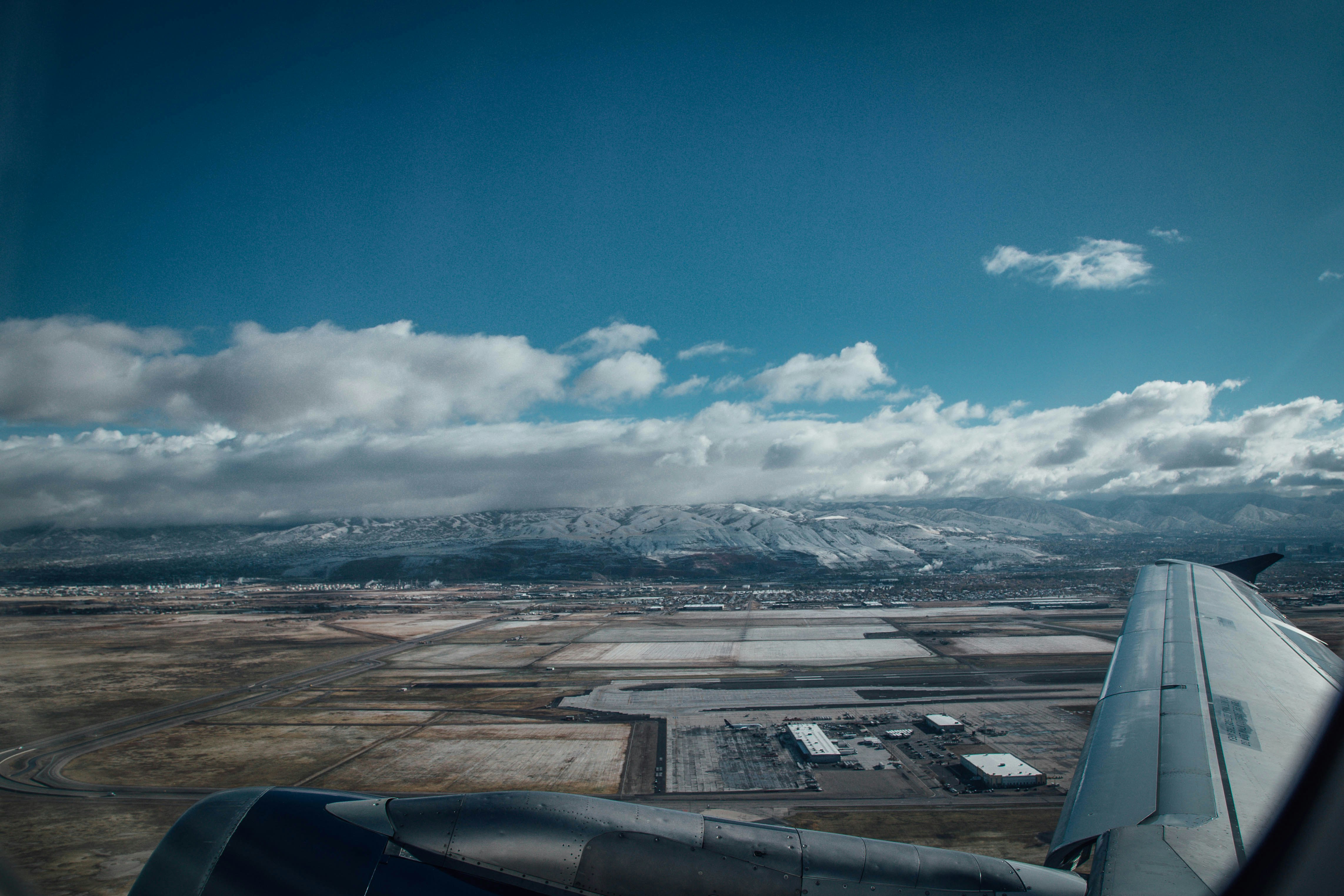 Airplane wing gliding above a vast landscape with snow-capped mountains and patches of green and brown fields.