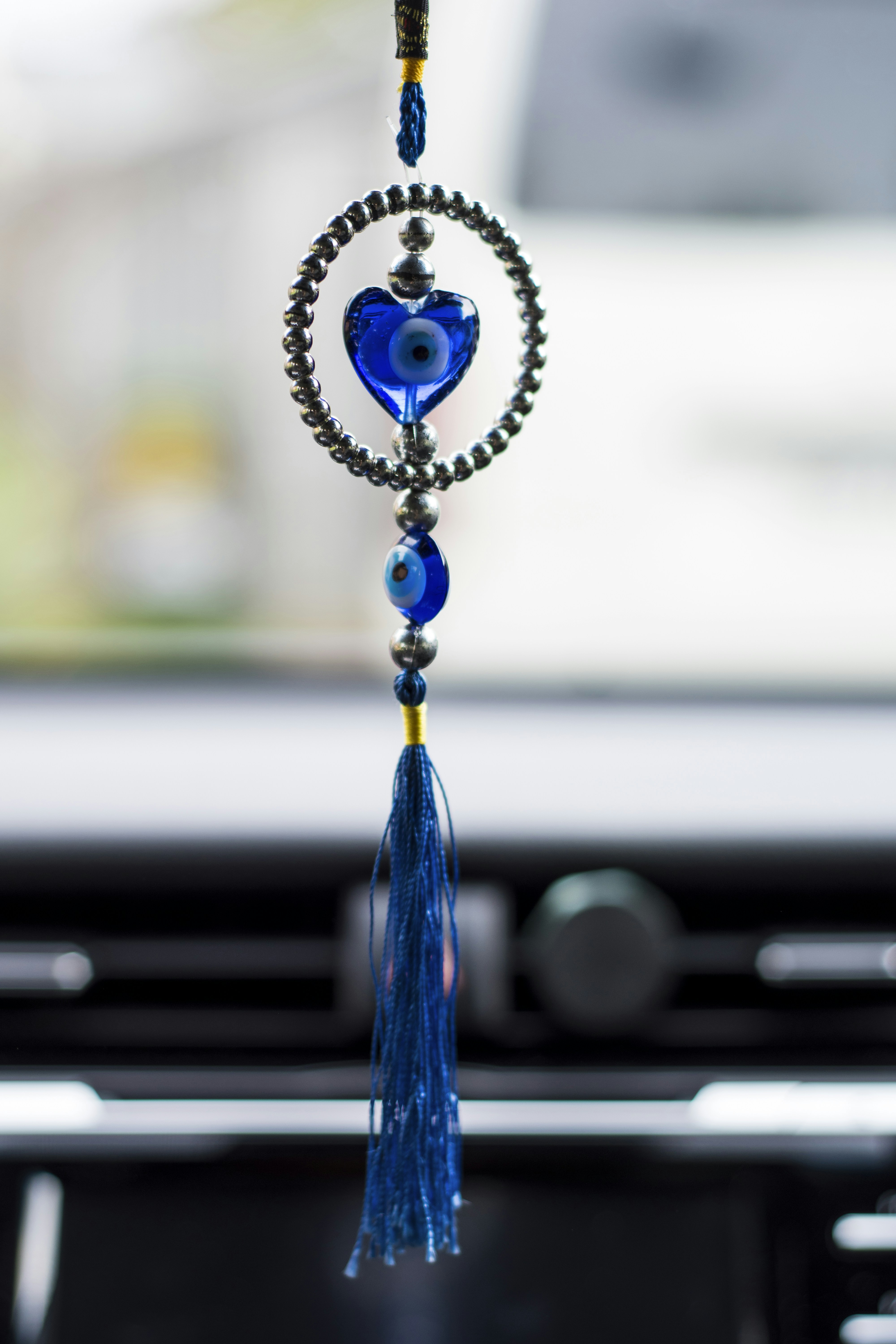 A blue glass charm featuring an eye and heart, suspended with silver beads, hangs from a tassel in a car interior. The charm symbolizes protection and good fortune.