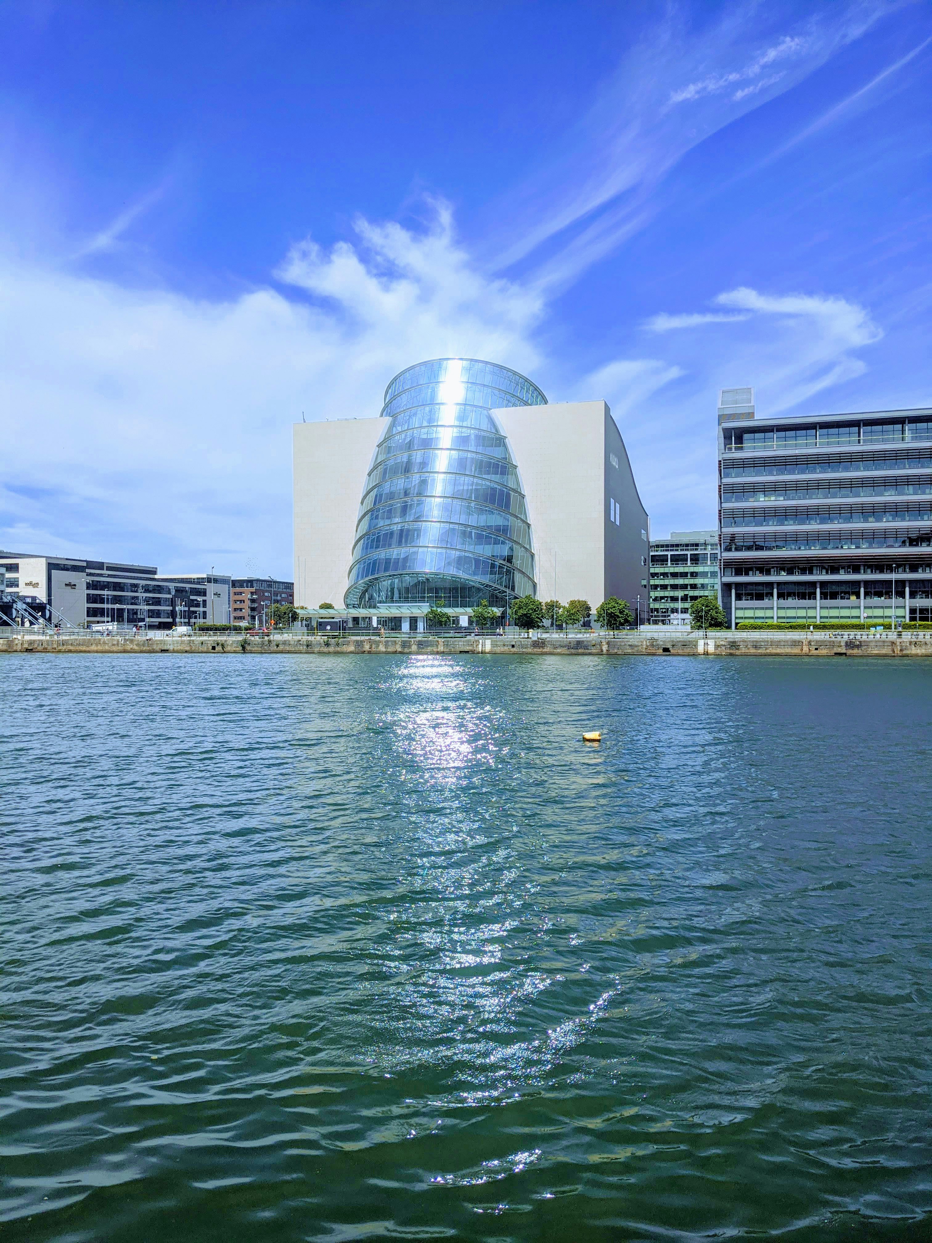 Curved glass tower rises along the riverfront beside modern office blocks, with sunlight glinting on the water.