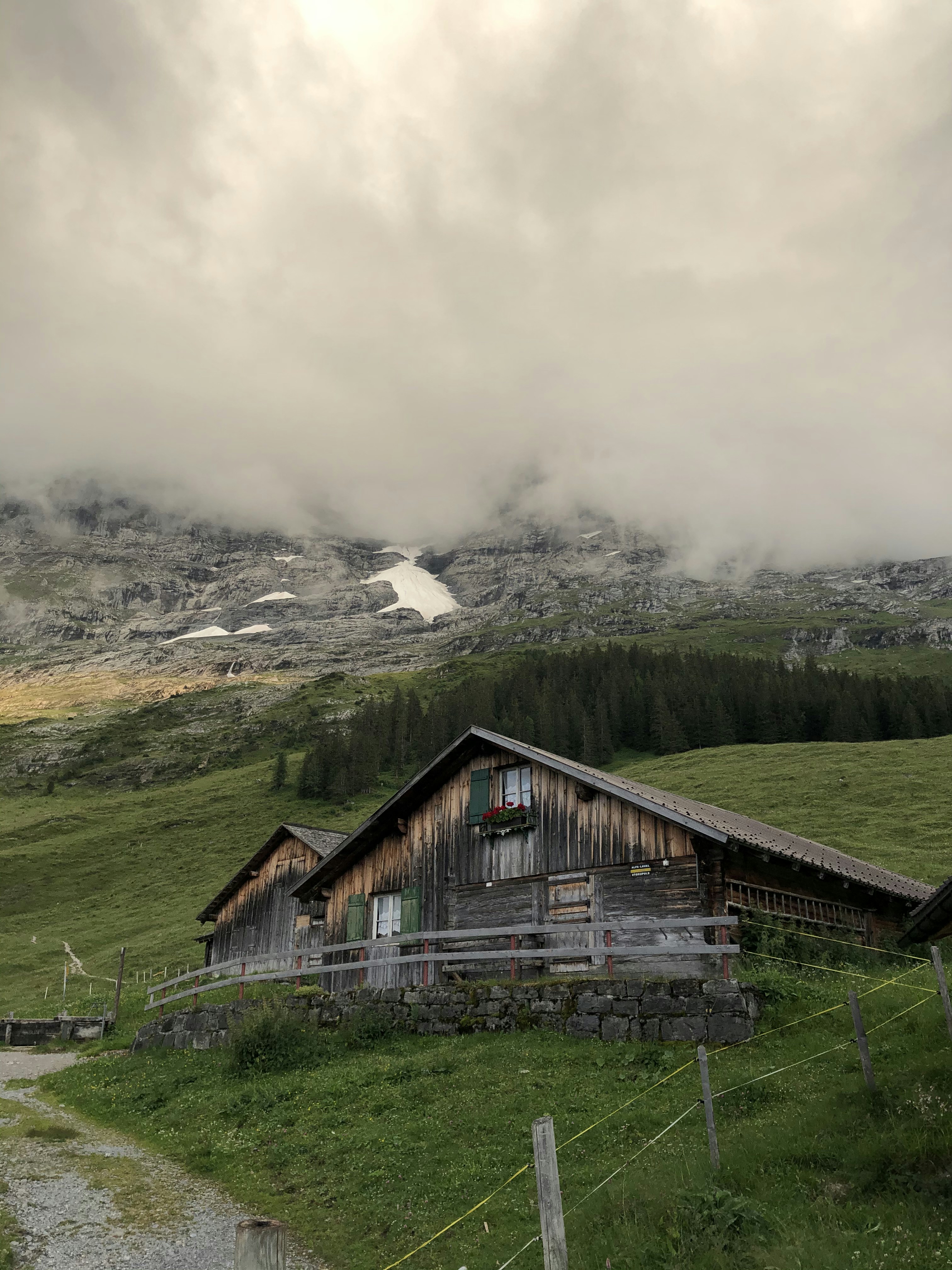 Rustic wooden cabin nestled in a verdant alpine landscape, with majestic mountains shrouded in clouds above. A serene blend of nature and architecture.