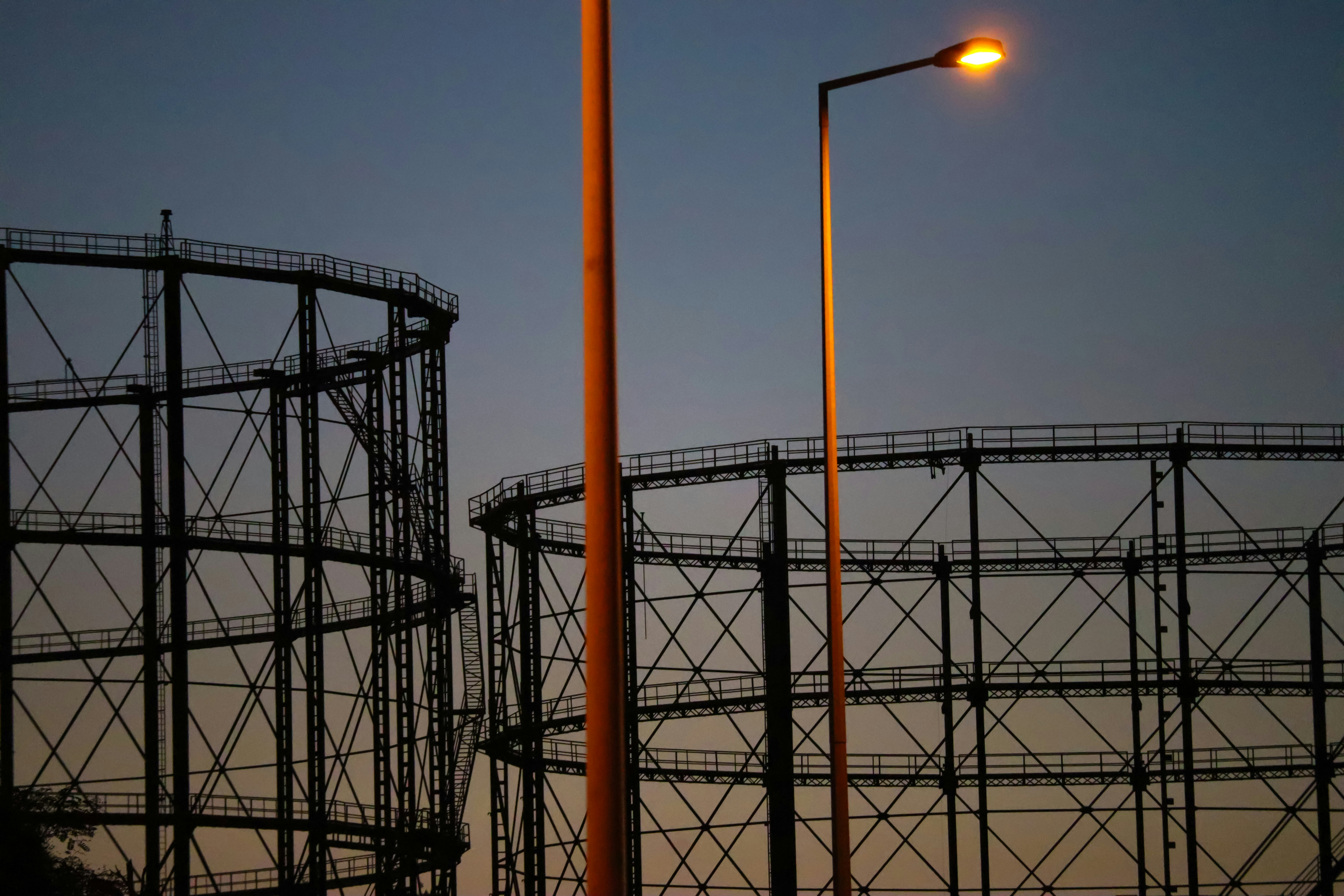 Dusk Over Gasometer Lattices: Twin Streetlamps Anchor a Skeletal Framework