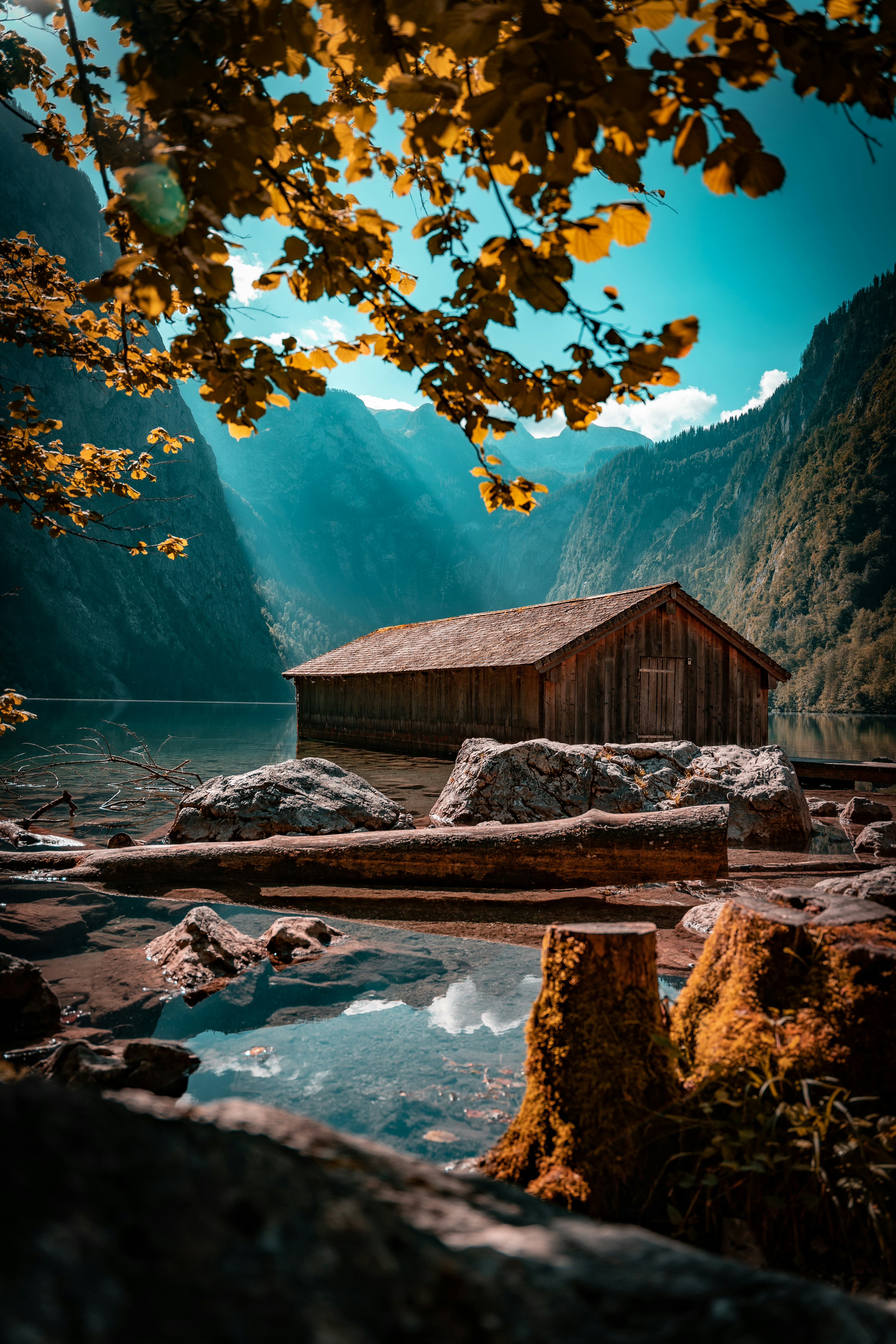 Wooden cabin nestled by a serene lake, surrounded by majestic mountains and vibrant autumn foliage. Reflections shimmer on the water's surface.