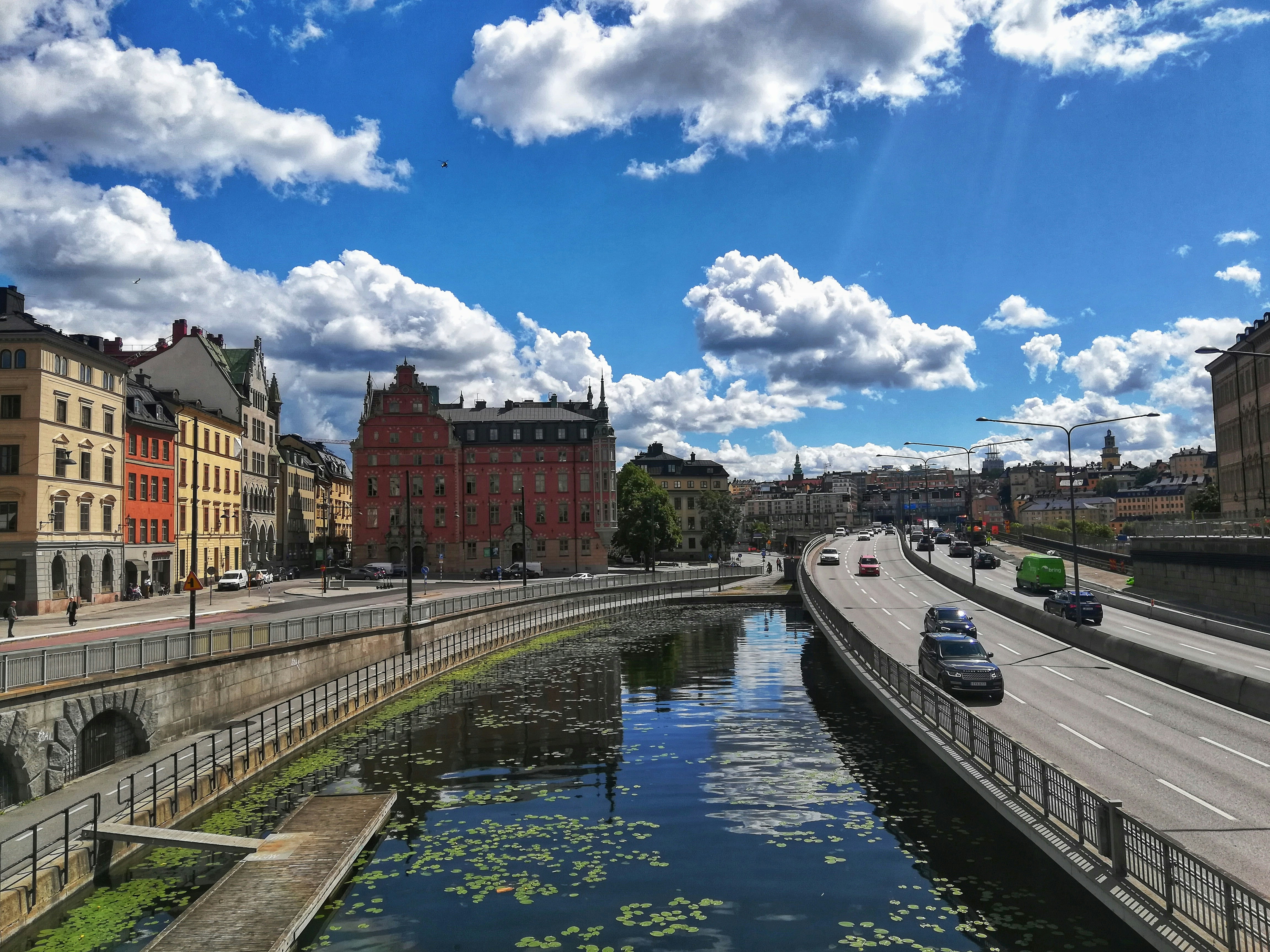 Colorful buildings lining a serene waterway under a bright blue sky with fluffy clouds. The scene captures urban life alongside nature.