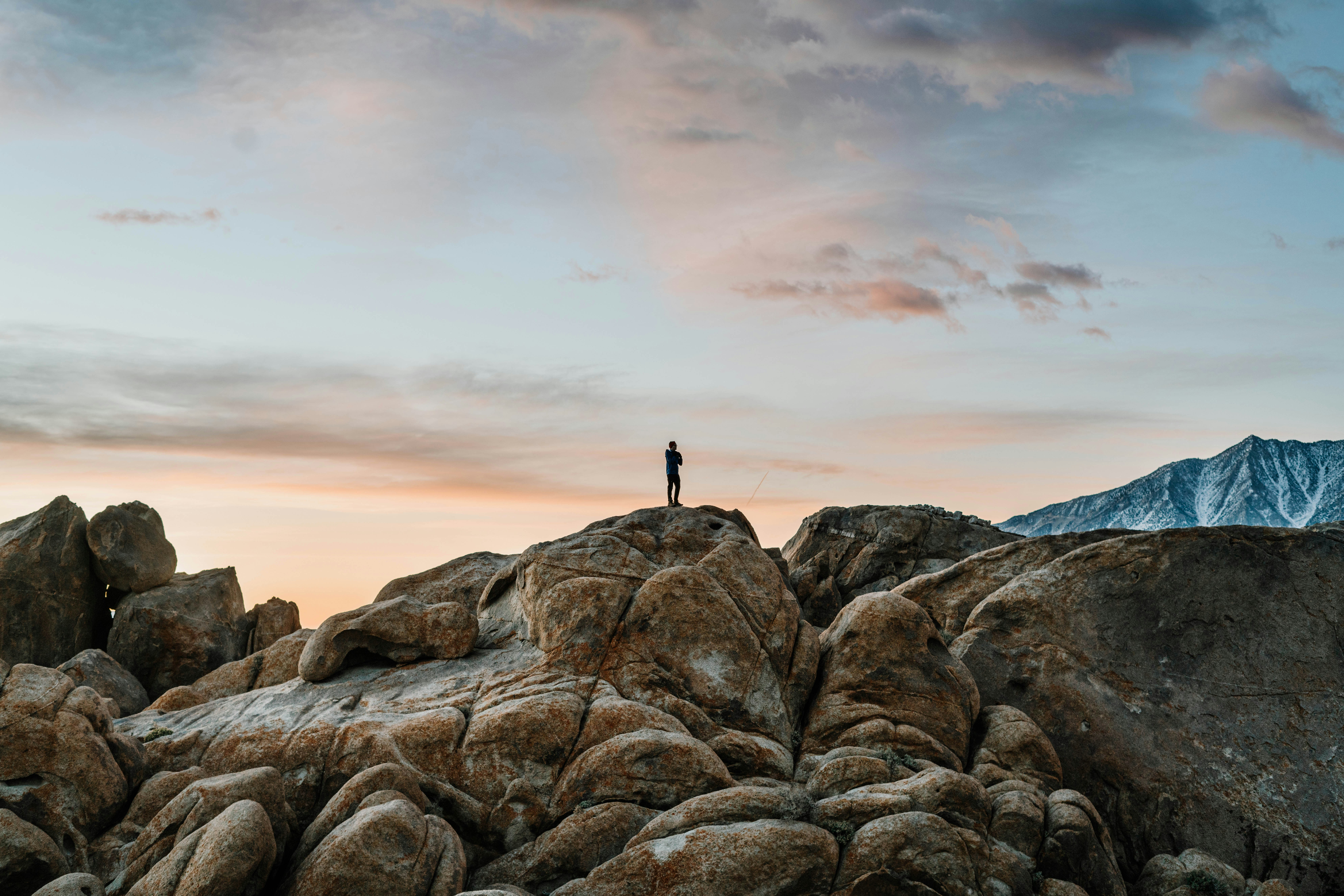Person standing atop rugged rock formation beneath a pastel sky during sunset.