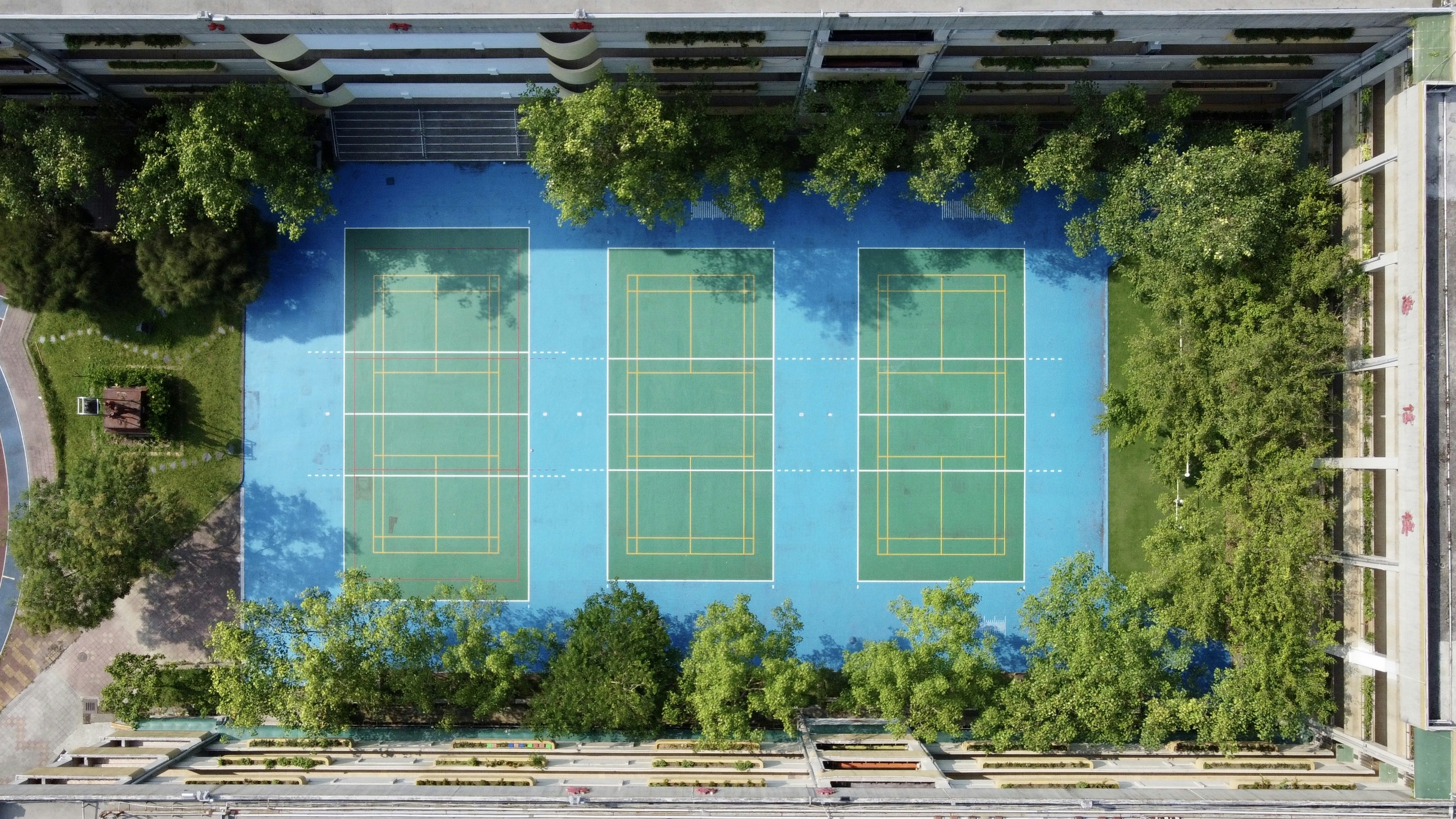 Aerial view of three tennis courts surrounded by lush greenery and modern architecture, showcasing a blend of recreational space and nature.