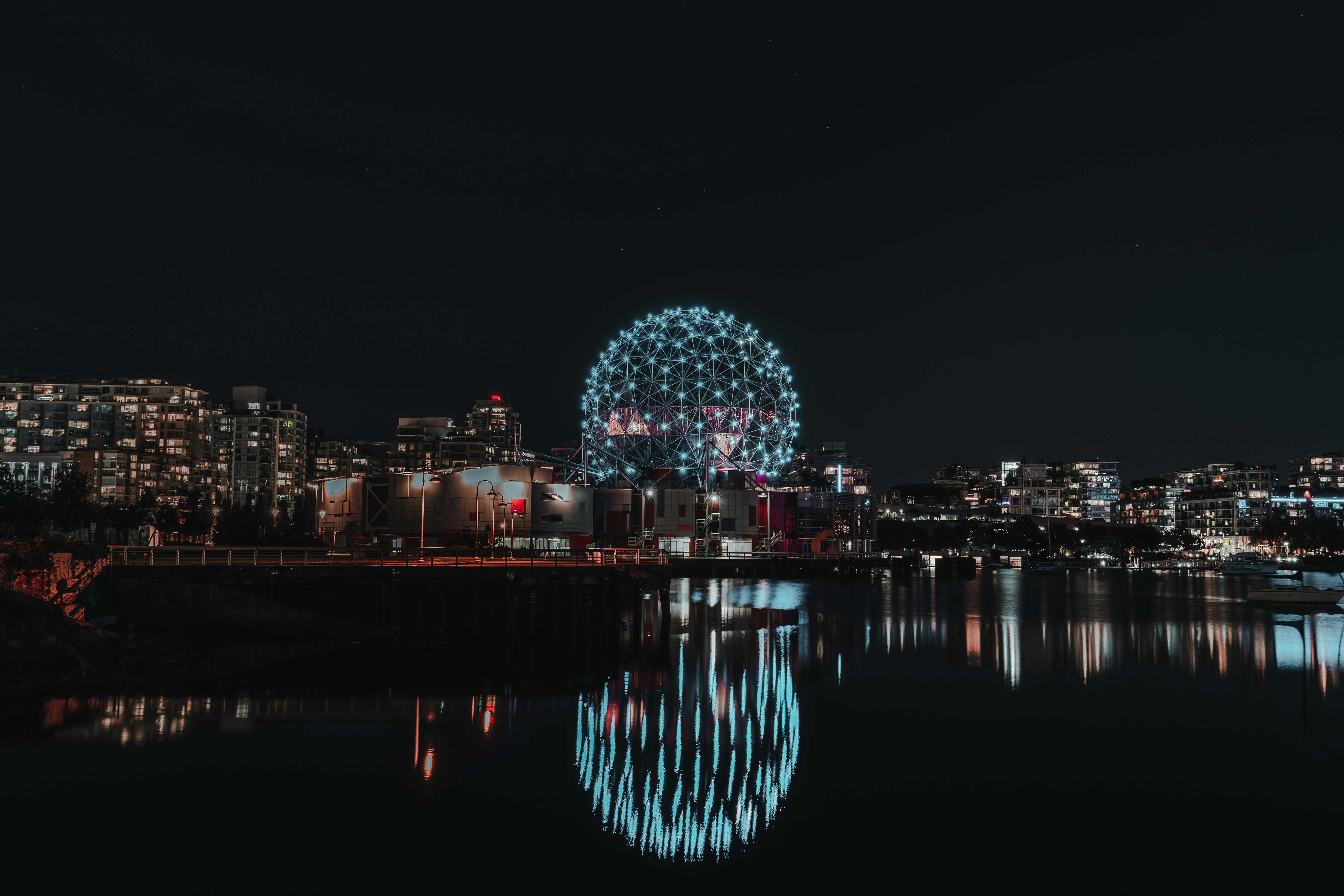 A vibrant geodesic dome shines against a dark sky, mirrored in the calm waters below. Urban lights create a striking contrast with the night backdrop.