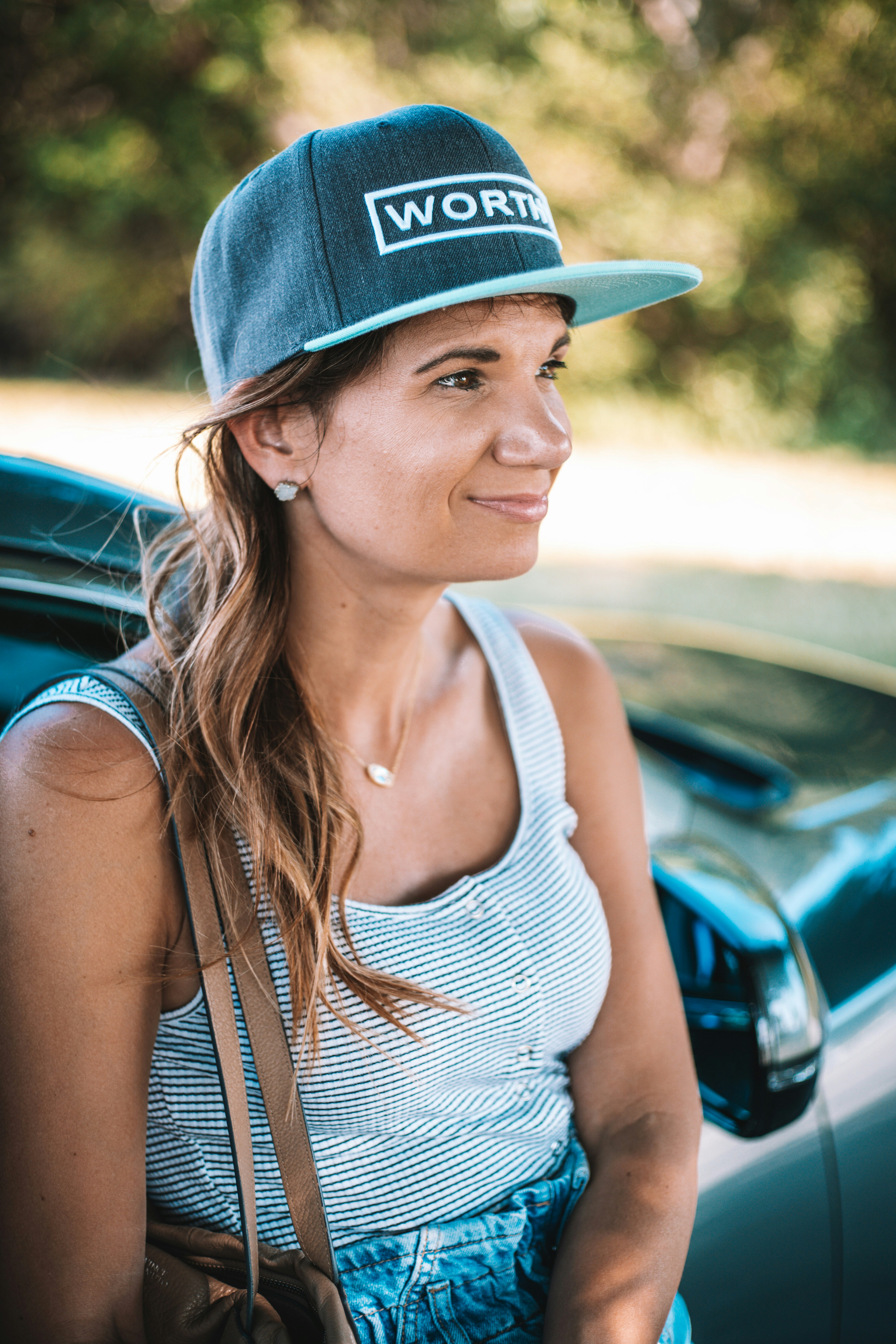 Woman wearing a cap with the word 'WORTHY' sits beside a car, exuding confidence and style. The background features soft greenery.
