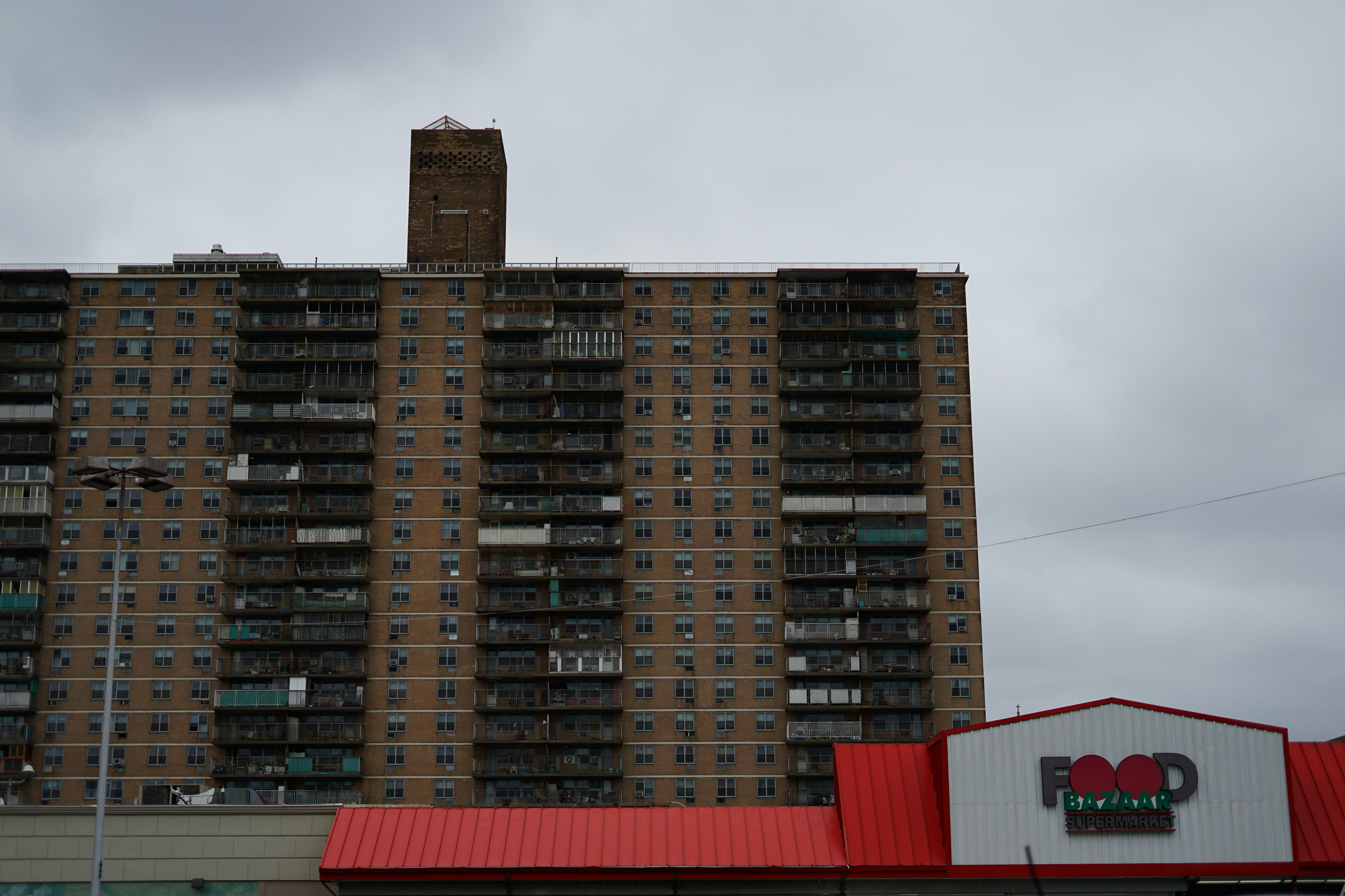 A weathered high-rise building showcases its aging balconies and windows, juxtaposed with a vibrant grocery store in the foreground. The scene reflects urban life and community dynamics.