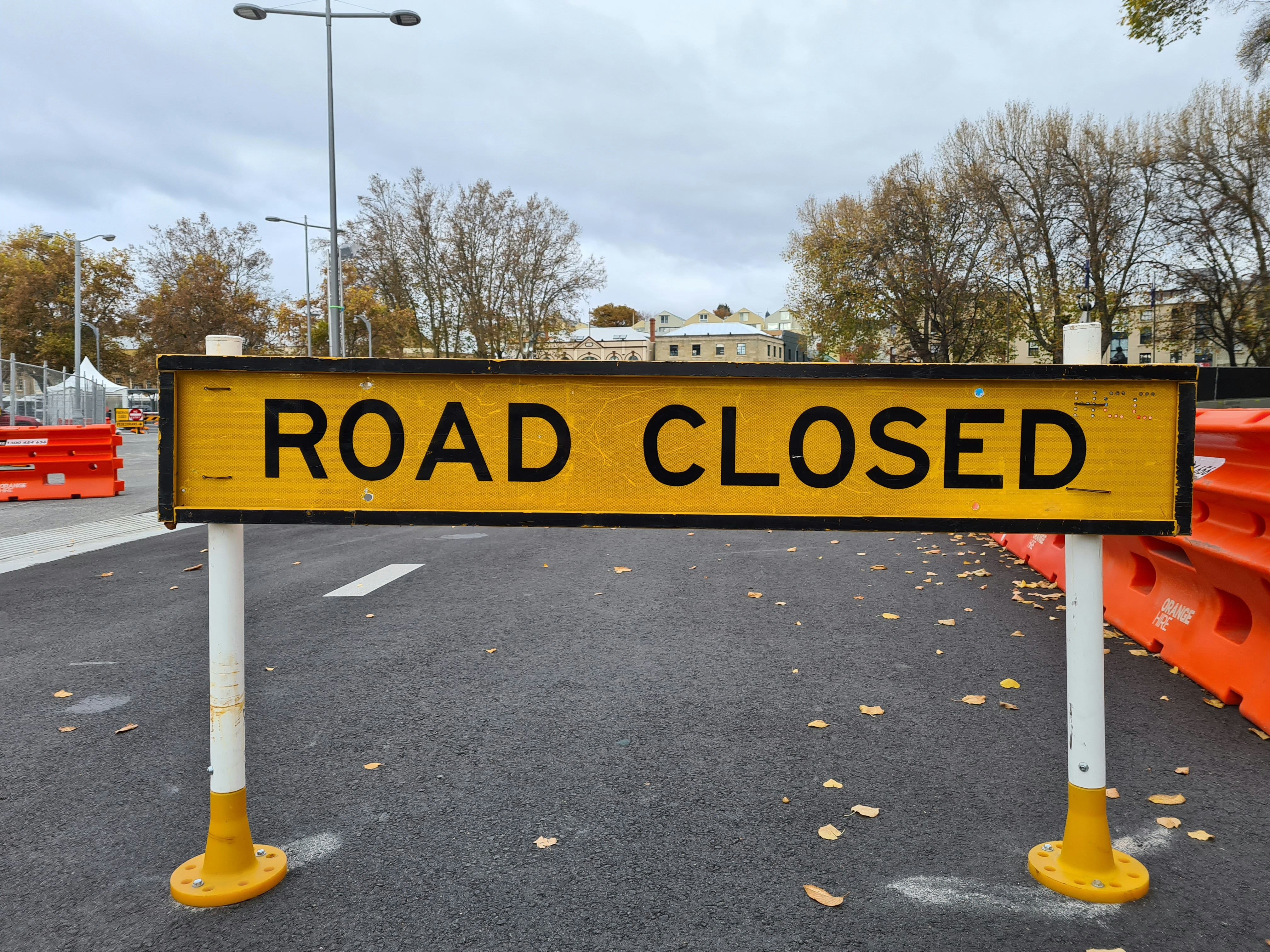 Yellow 'Road Closed' sign on freshly paved street with orange barriers and autumn trees.