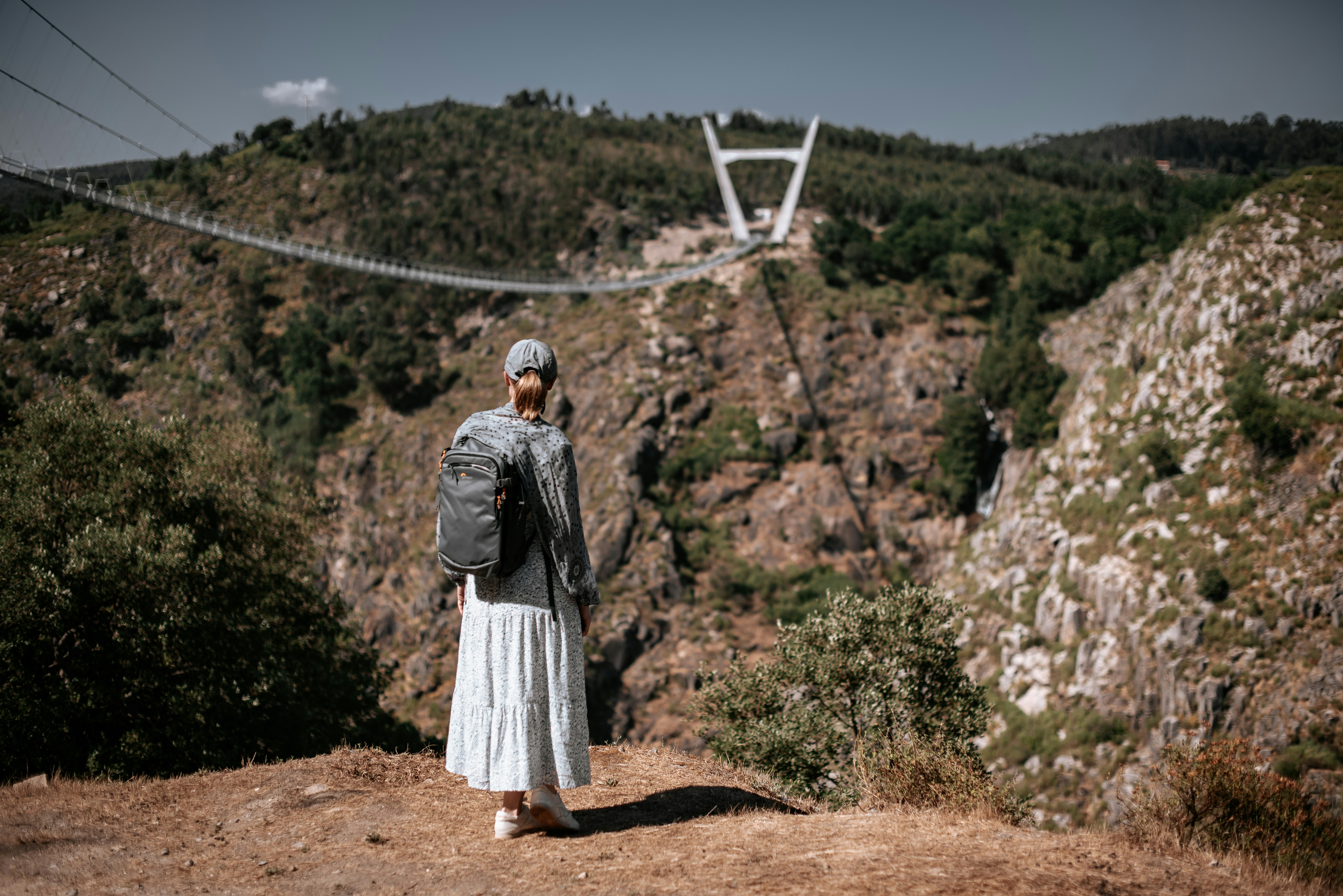 A lone traveler gazes at a striking suspension bridge spanning a deep gorge, surrounded by lush greenery and rocky terrain.