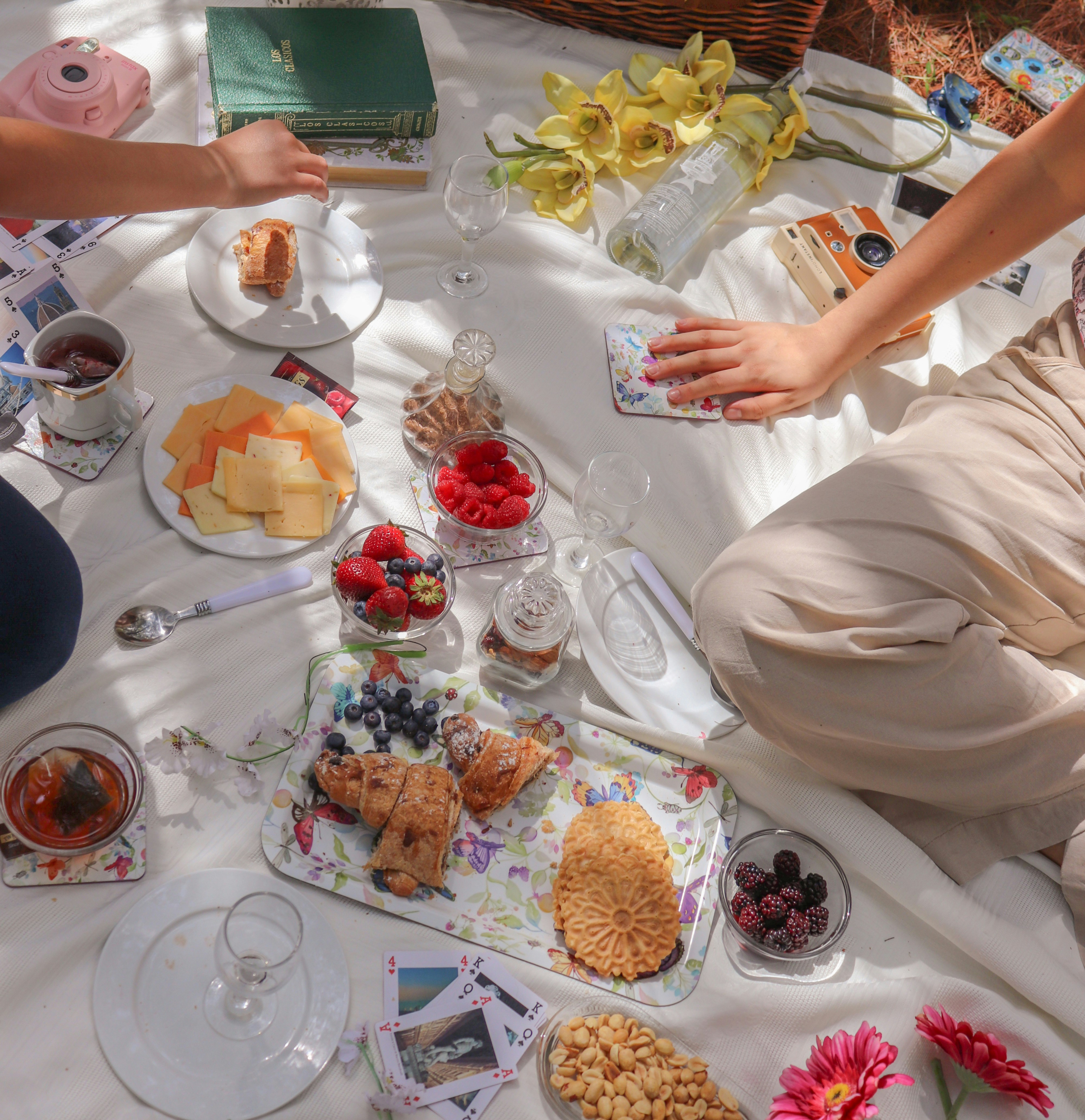 Sunlit picnic spread on a white cloth features berries, pastries, cheese, and flowers arranged around glassware. This top-down photograph captures a casual, convivial moment of sharing.