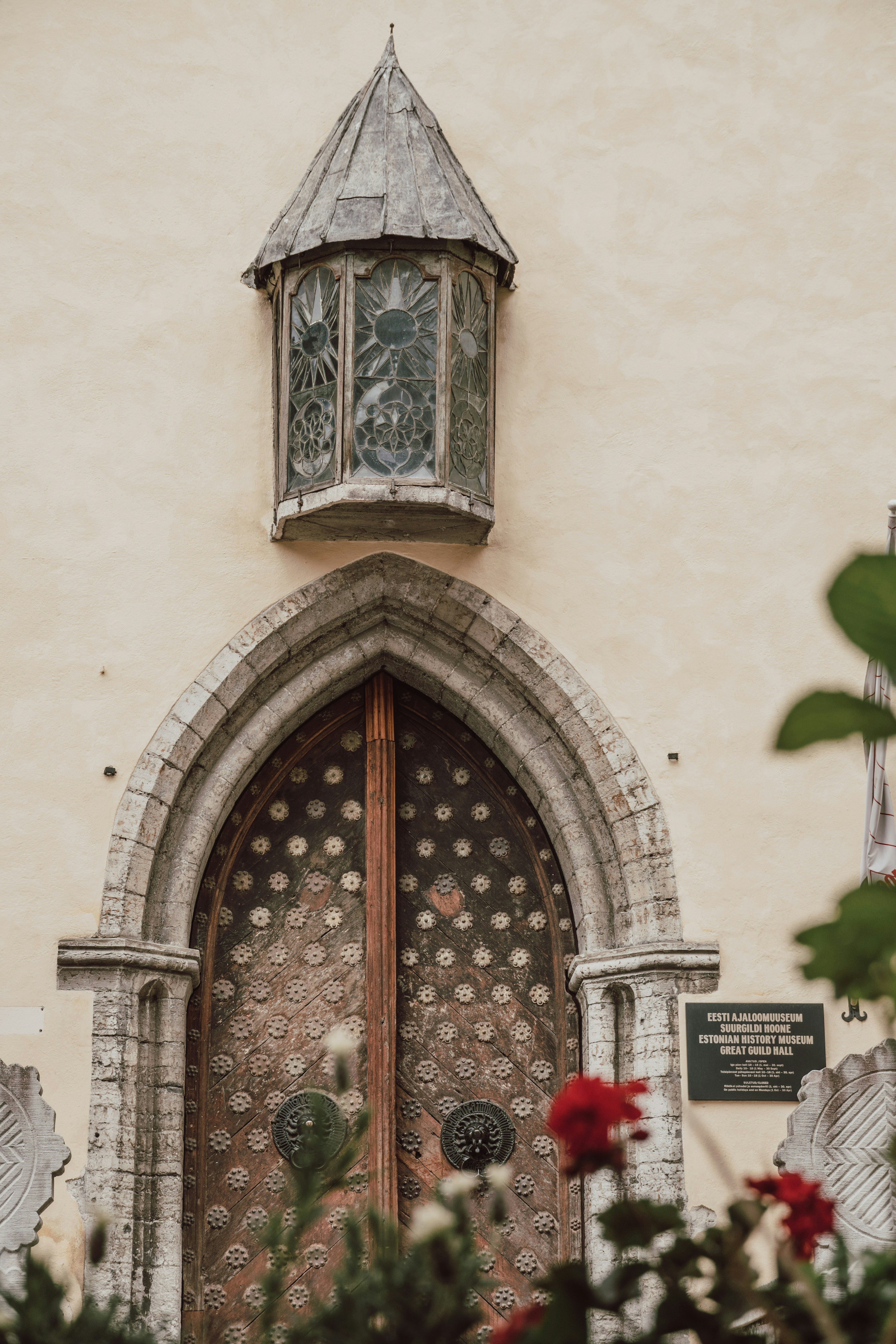 Intricate wooden door adorned with floral patterns, framed by a stone arch and topped with a decorative window. A plaque nearby provides historical context.