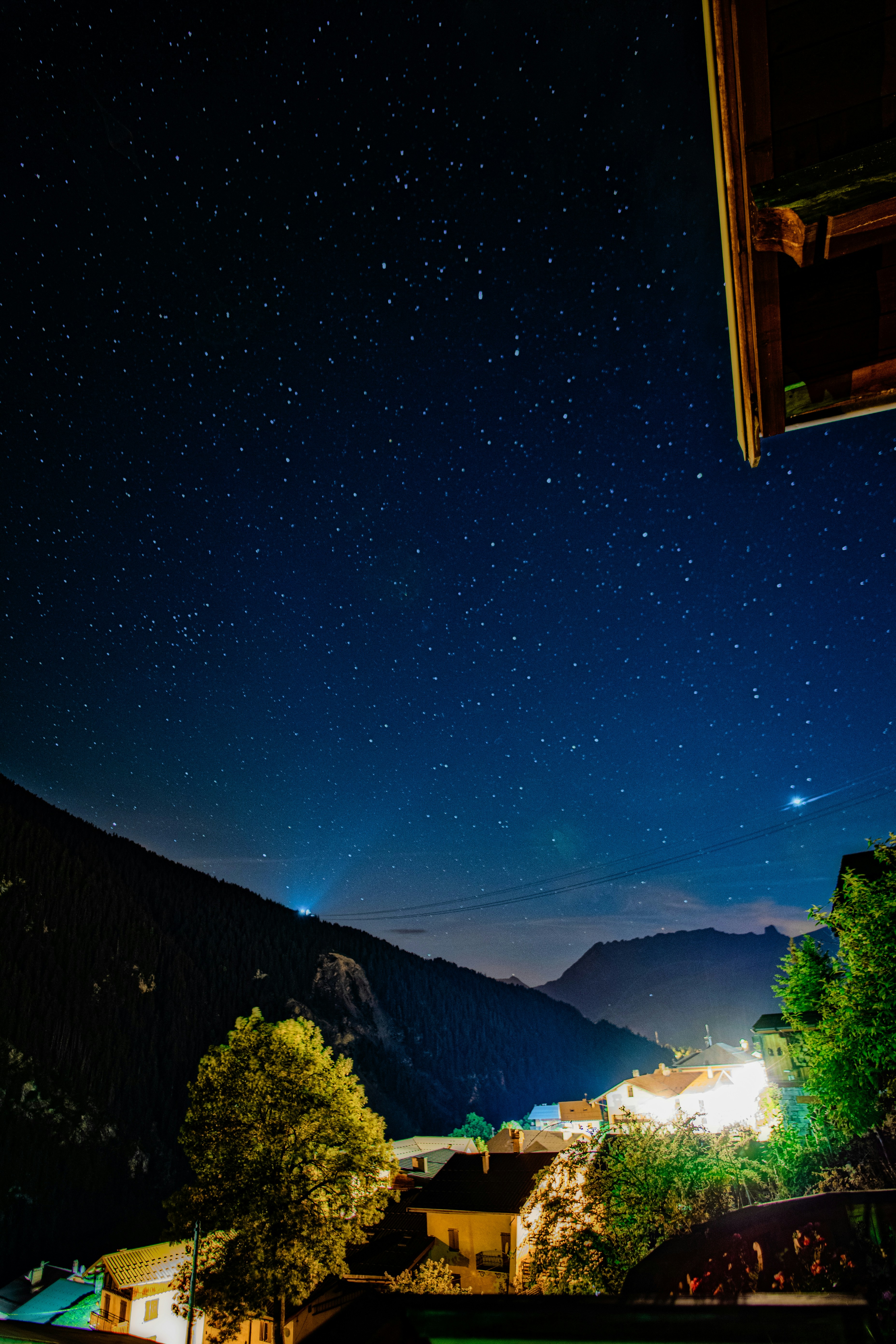 Star-filled night sky over a mountain village, with glowing lights and silhouetted peaks.