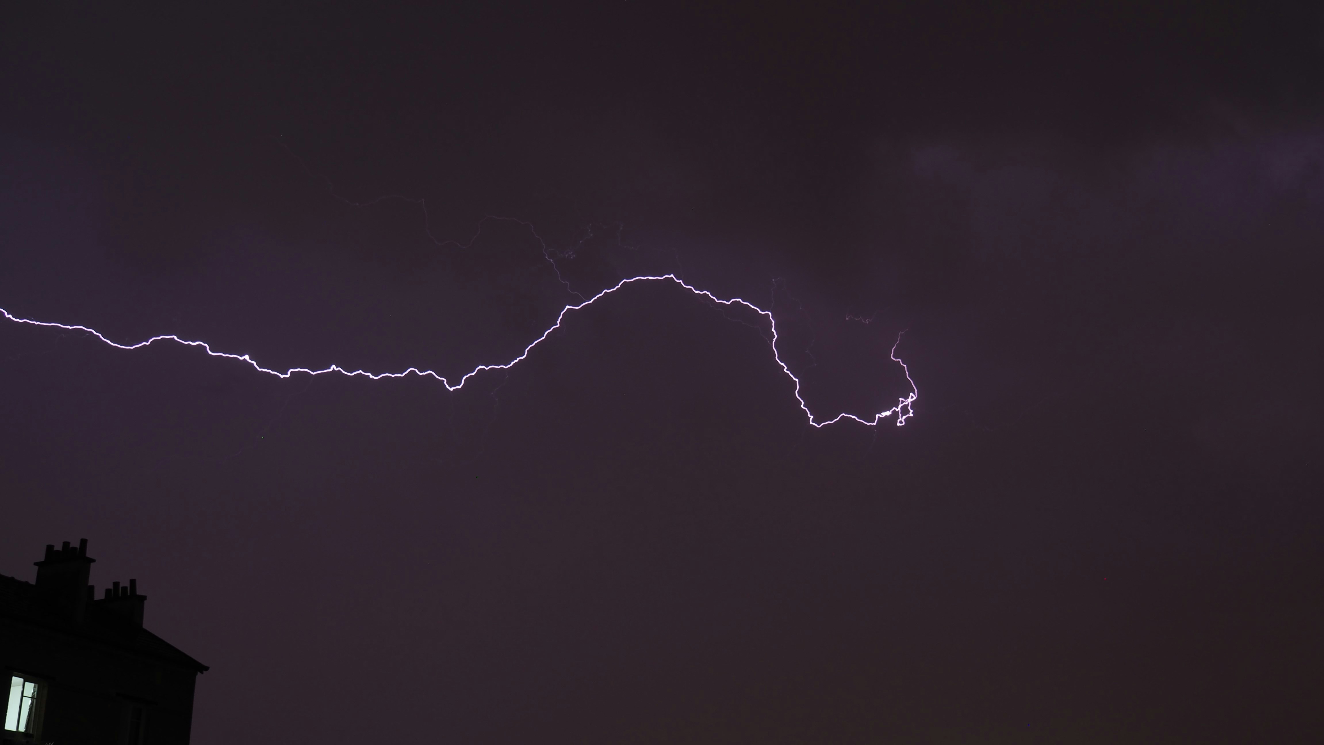 Dramatic lightning streaks across a darkened sky, illuminating the clouds with a vivid glow. The silhouette of a building frames the bottom of the scene.