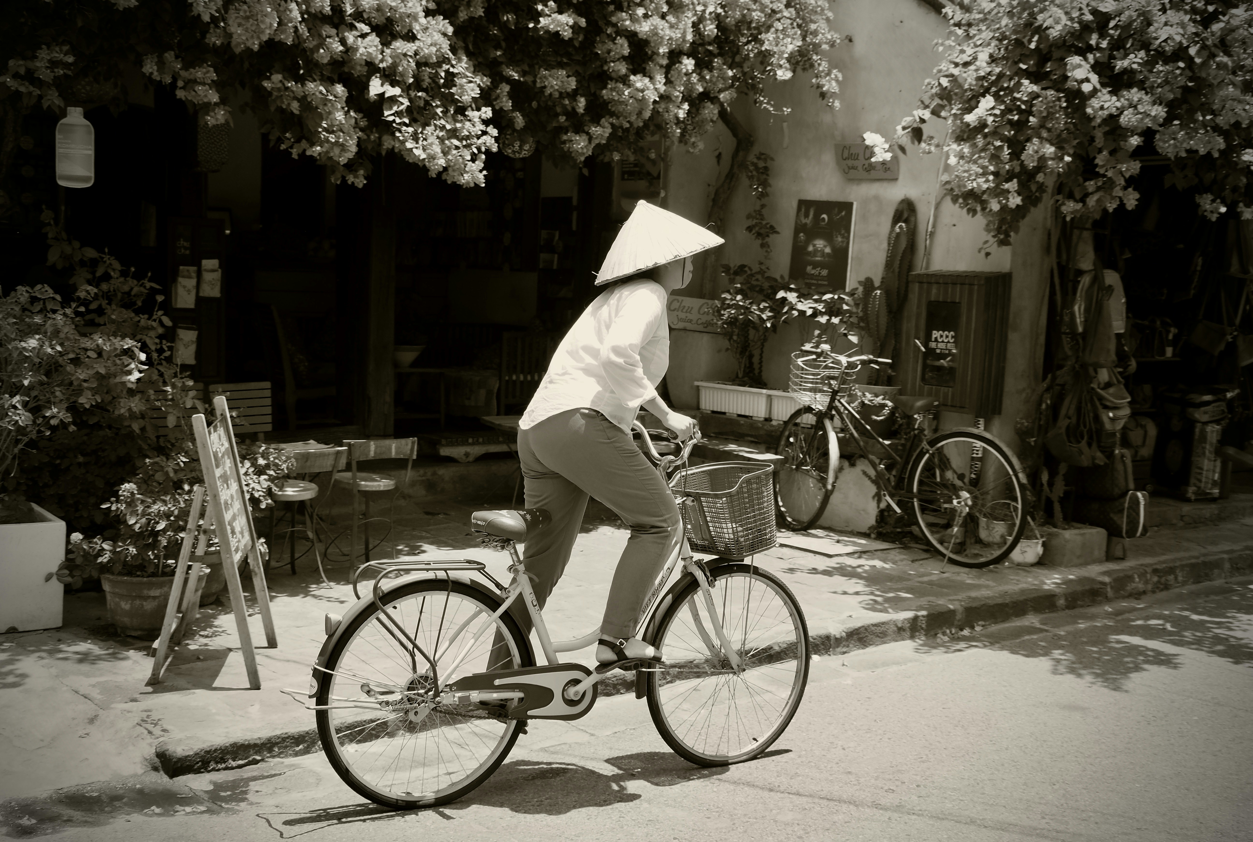 A person in traditional attire rides a bicycle past quaint storefronts adorned with blooming flora.