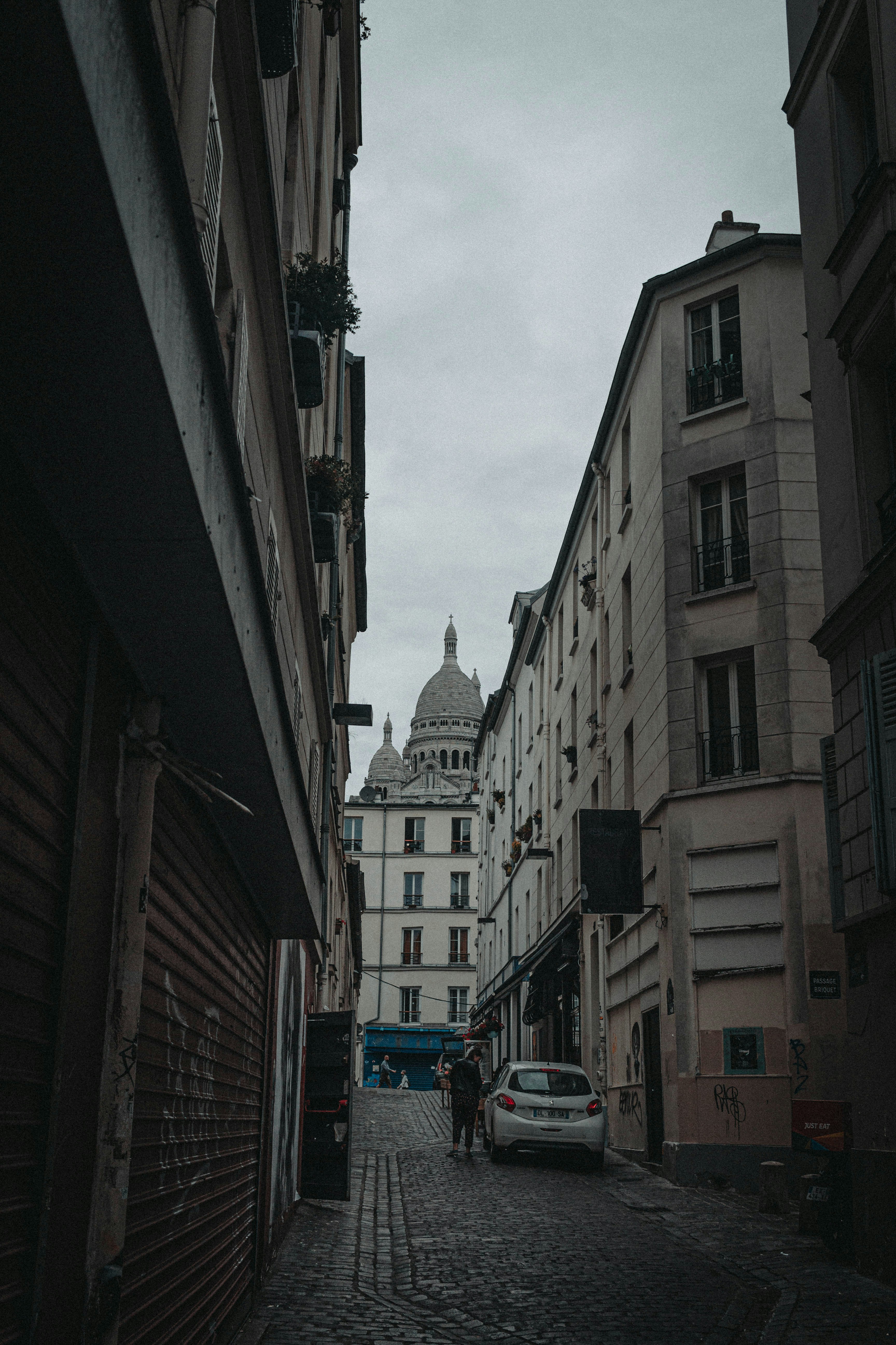 Narrow cobblestone street leading up to the iconic dome of Sacré-Cœur Basilica in Paris, framed by quaint buildings. A couple strolls down the path.