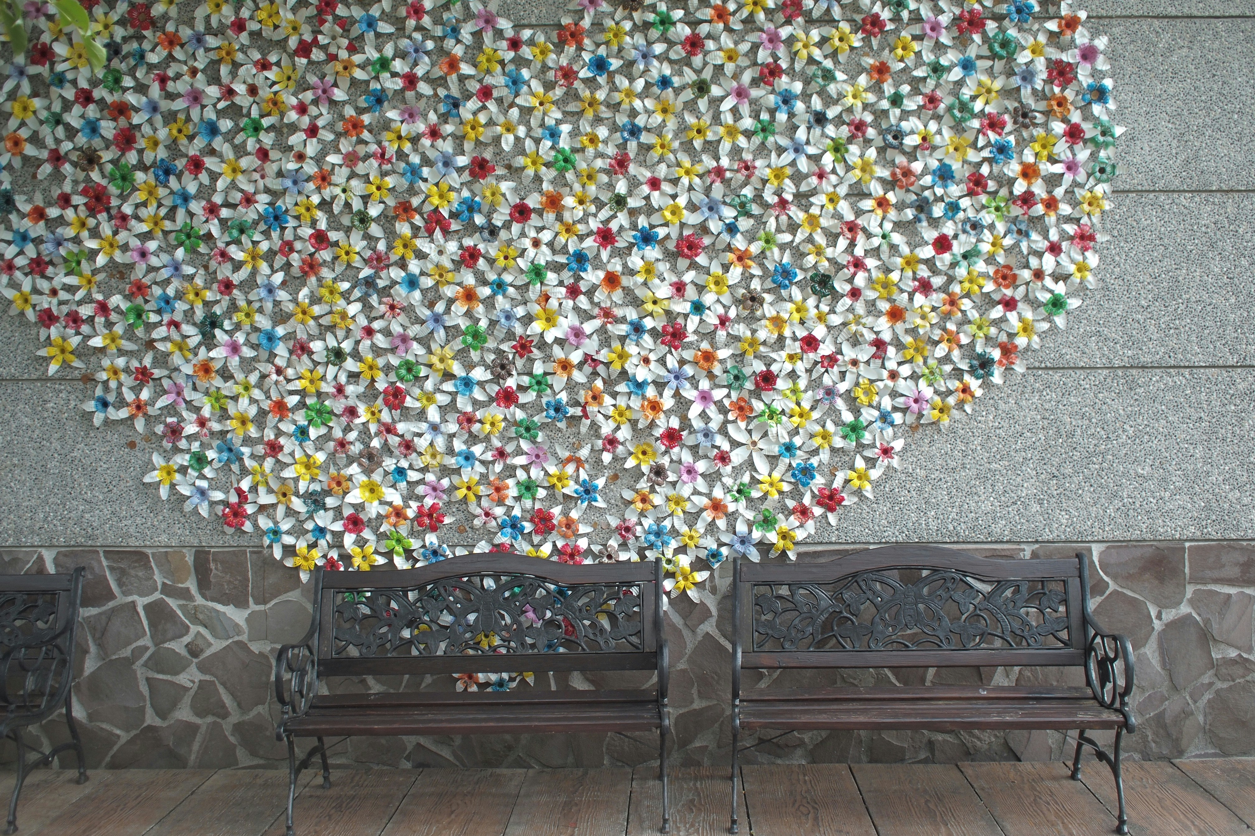 Colorful paper flowers arranged in a heart shape on a textured wall, with two empty benches below inviting rest and reflection.