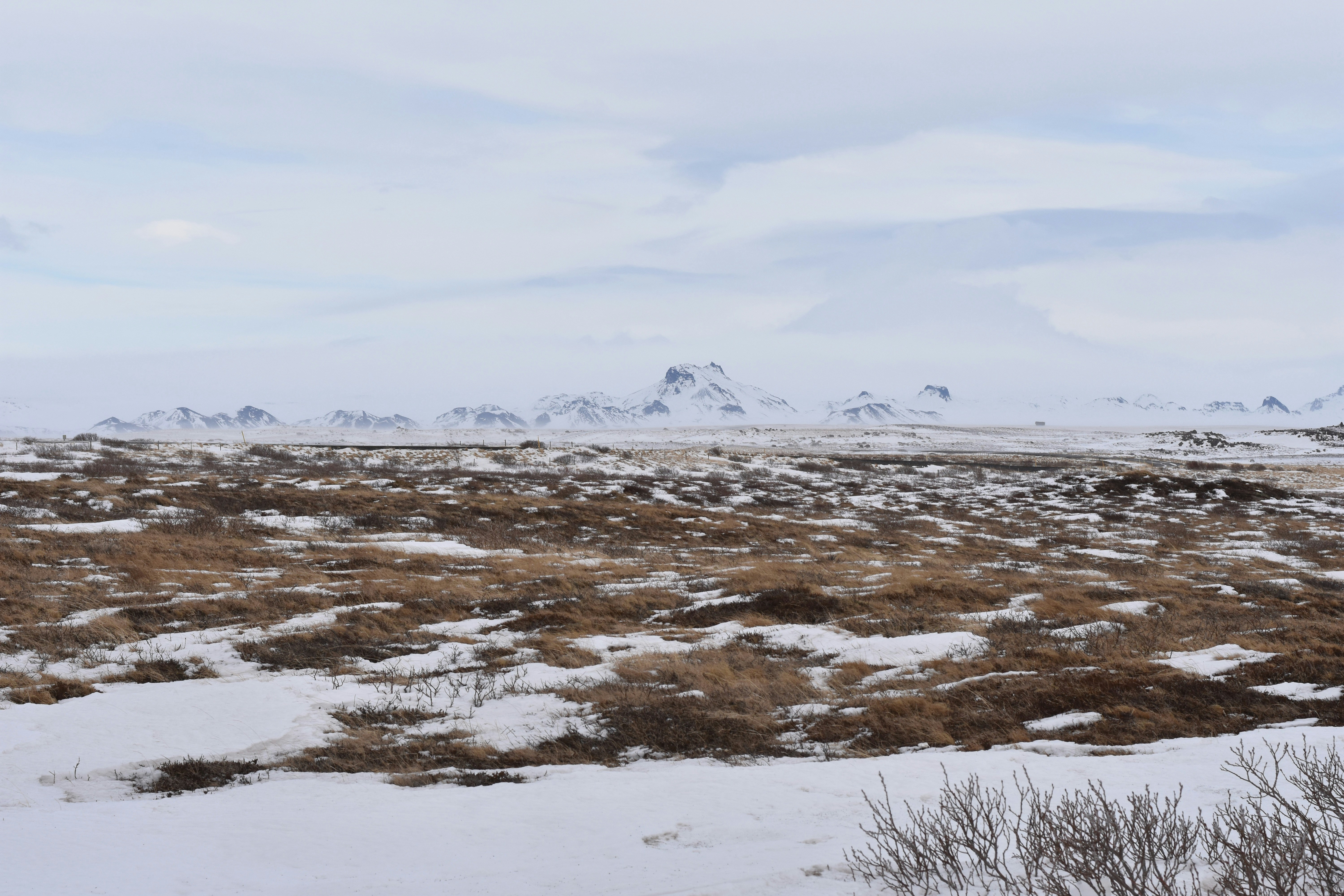 Expansive snow-covered landscape with patches of brown grass under a cloudy sky, hinting at distant mountains shrouded in mist.