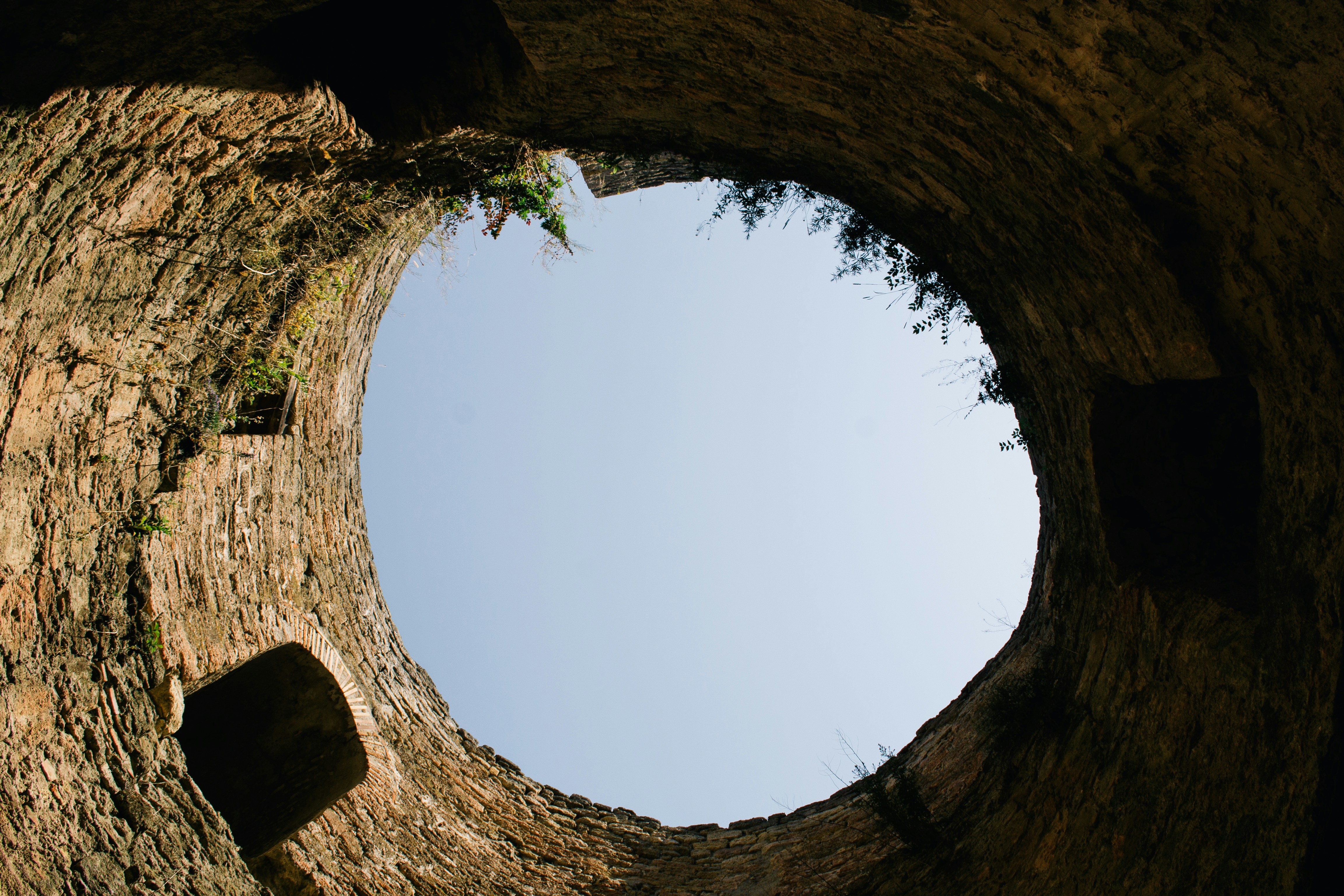 View looking up through a circular stone structure, framed by rugged walls and greenery, revealing a clear blue sky.