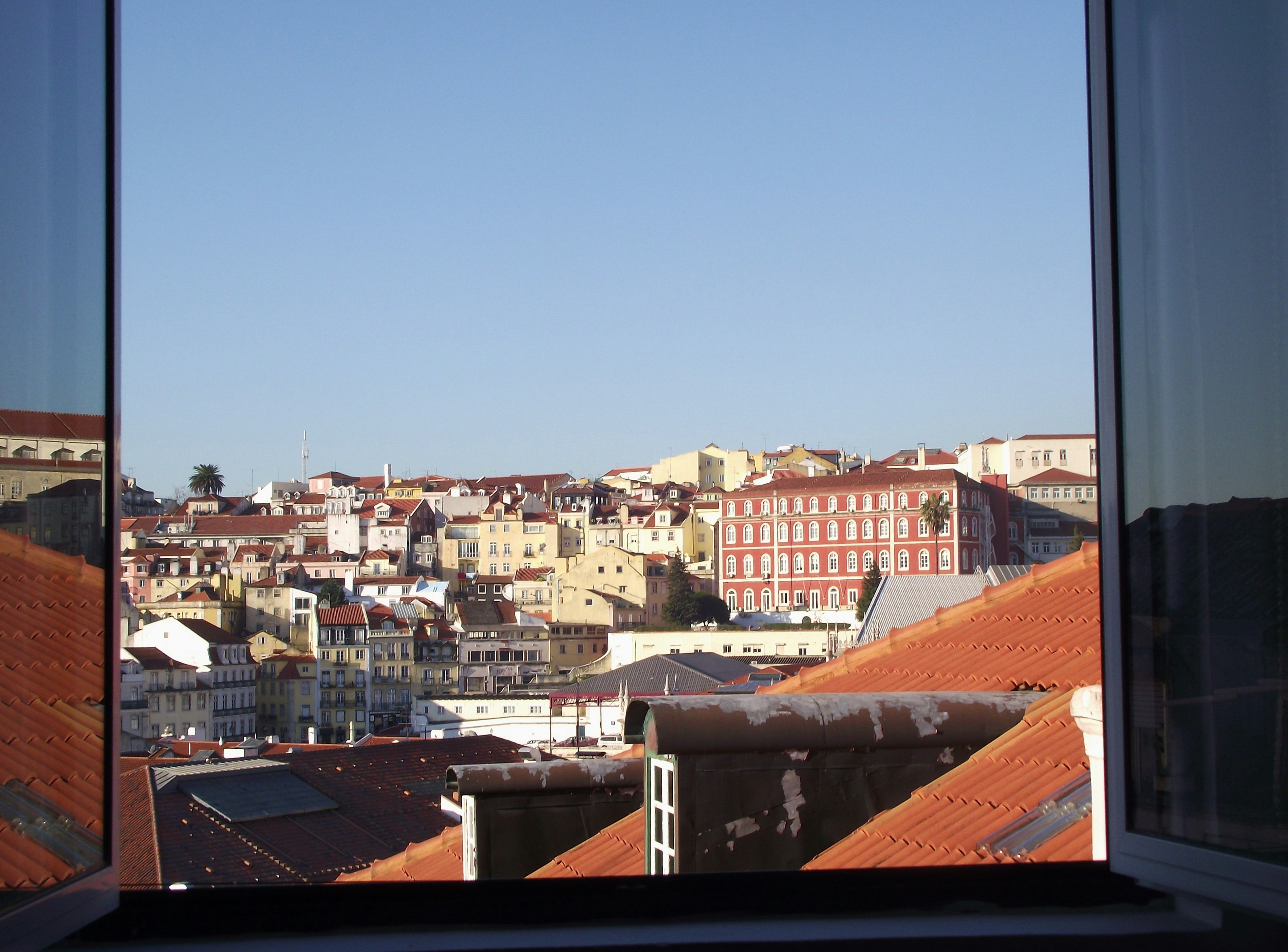 Colorful hillside cityscape framed by dark window panes, with red-orange tiled rooftops in the foreground and pastel buildings stretching toward a clear blue sky.
