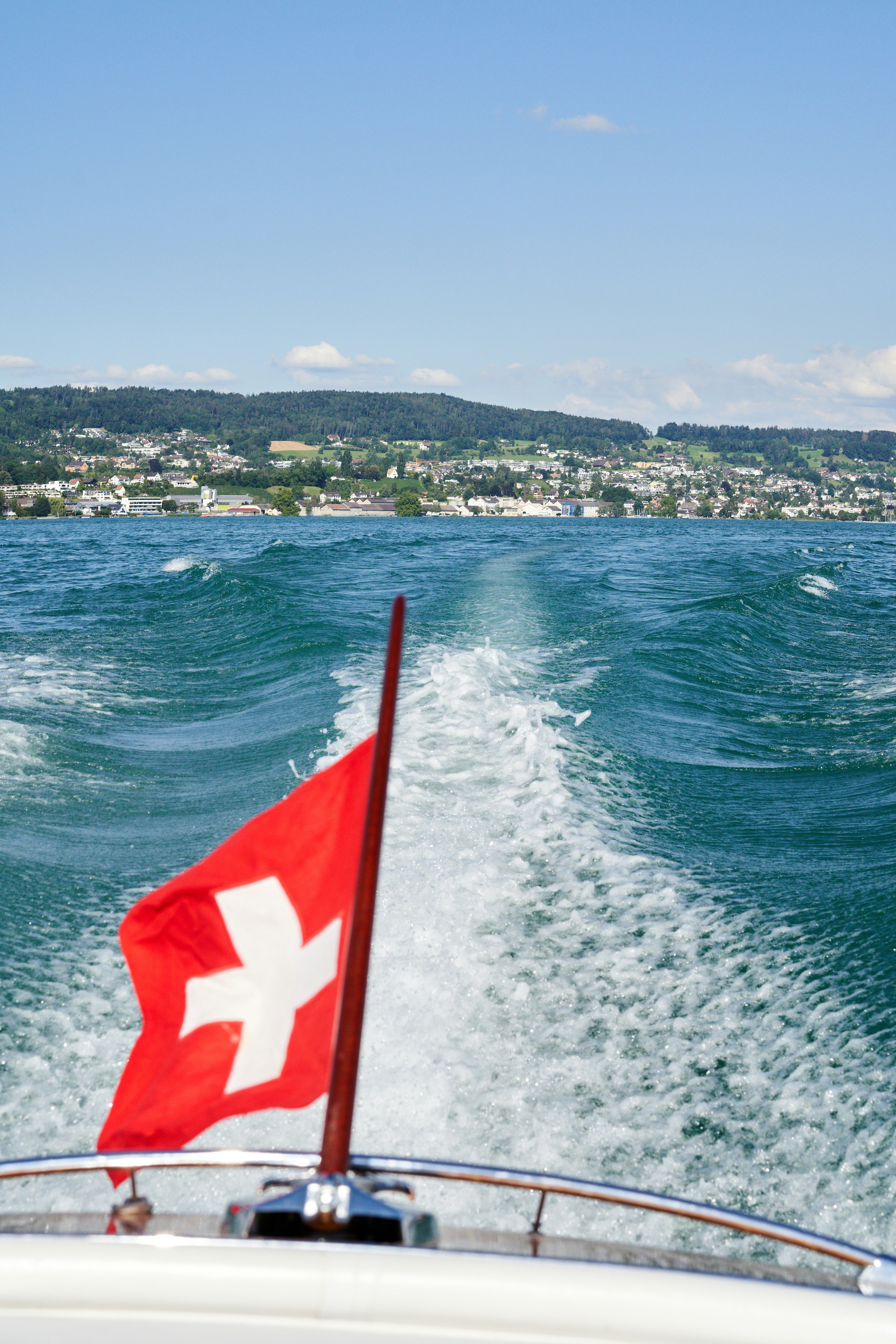 Swiss flag waves proudly as a boat cuts through tranquil waters, revealing a picturesque lakeside town in the background.