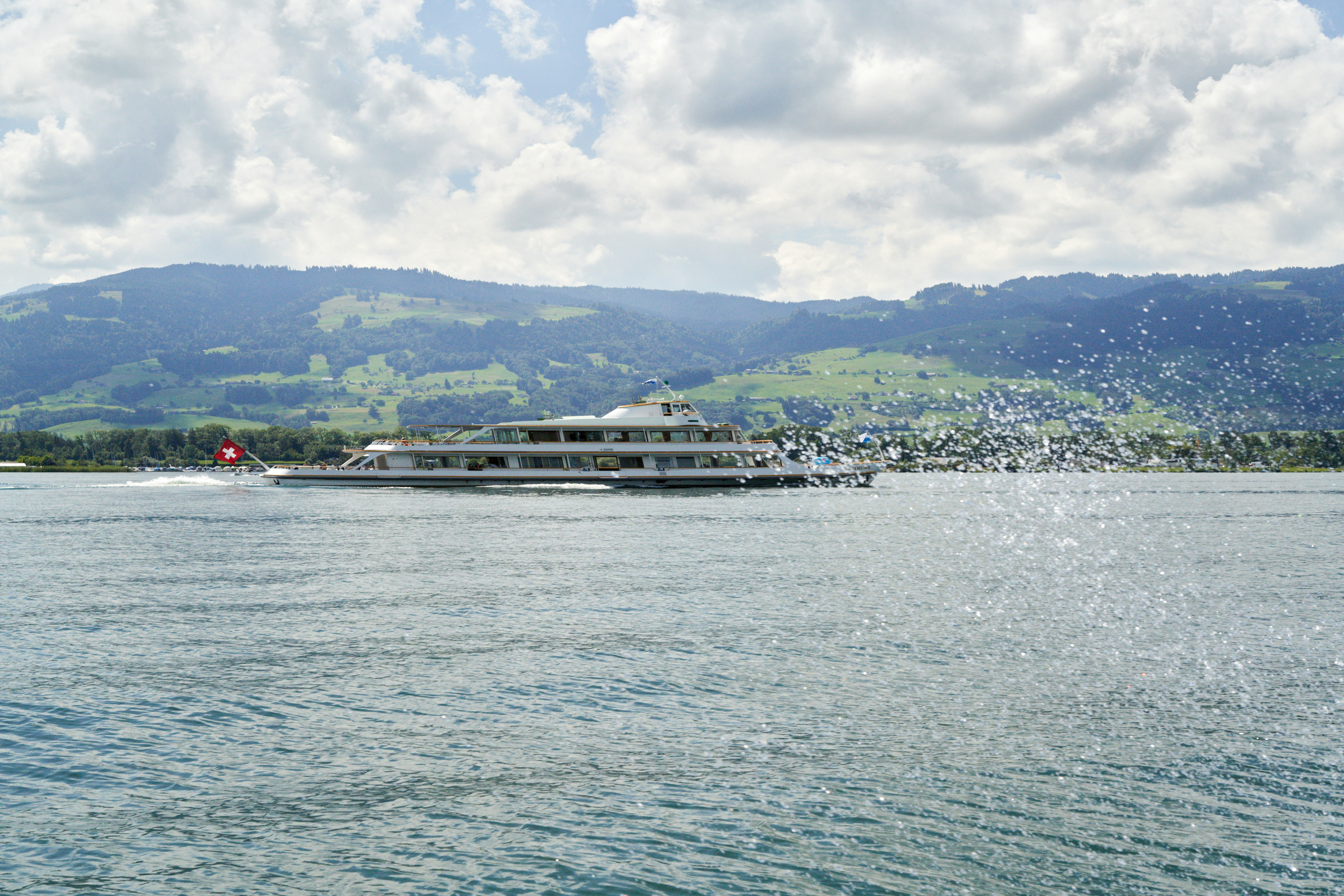Wake Across a Swiss Lake: A Sleek Yacht Slices Past Green Hills Under a Cloudy Sky