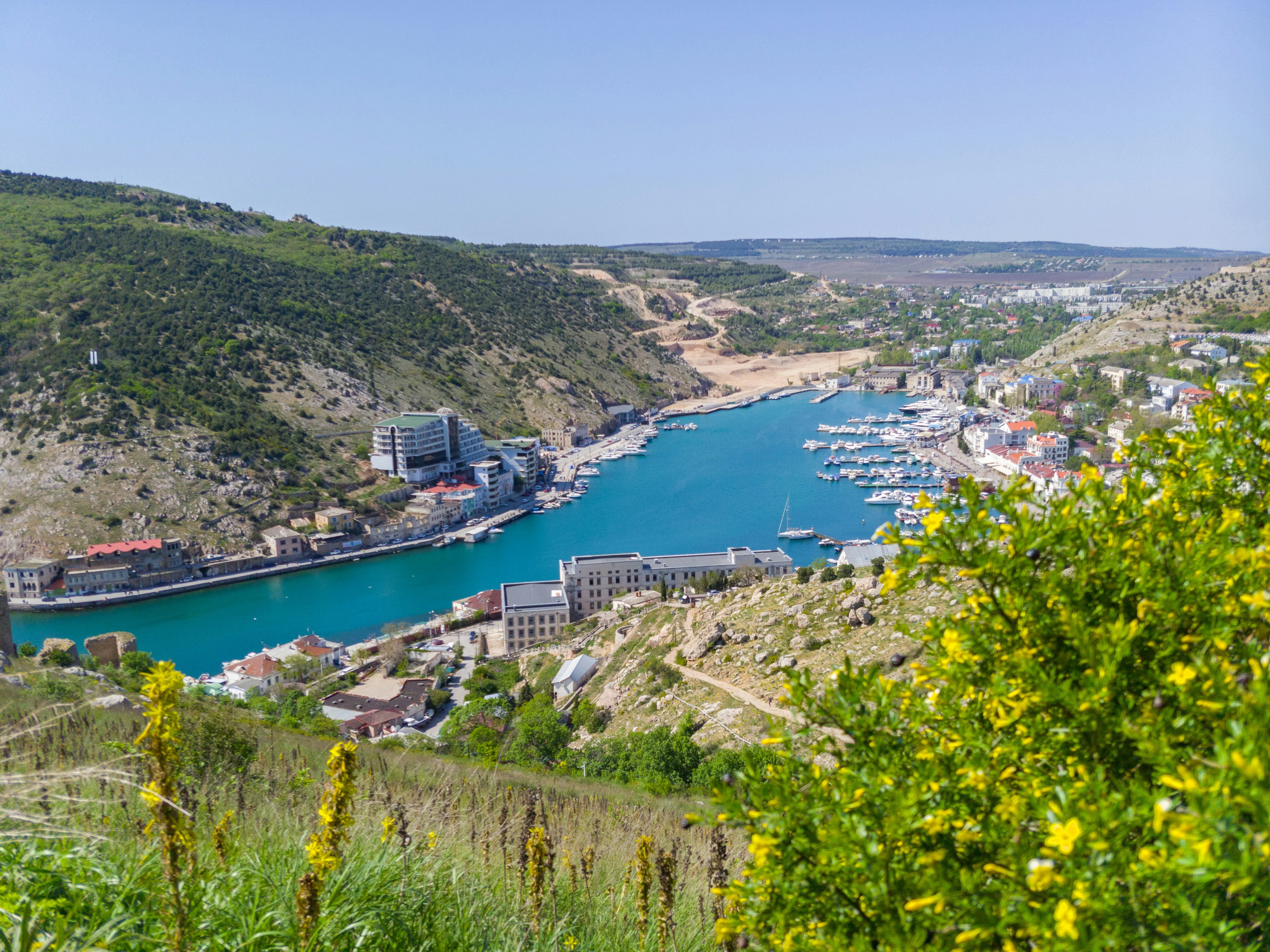 Vibrant marina nestled between lush hills, showcasing boats and colorful buildings along the waterfront. The scene reflects a tranquil coastal lifestyle.