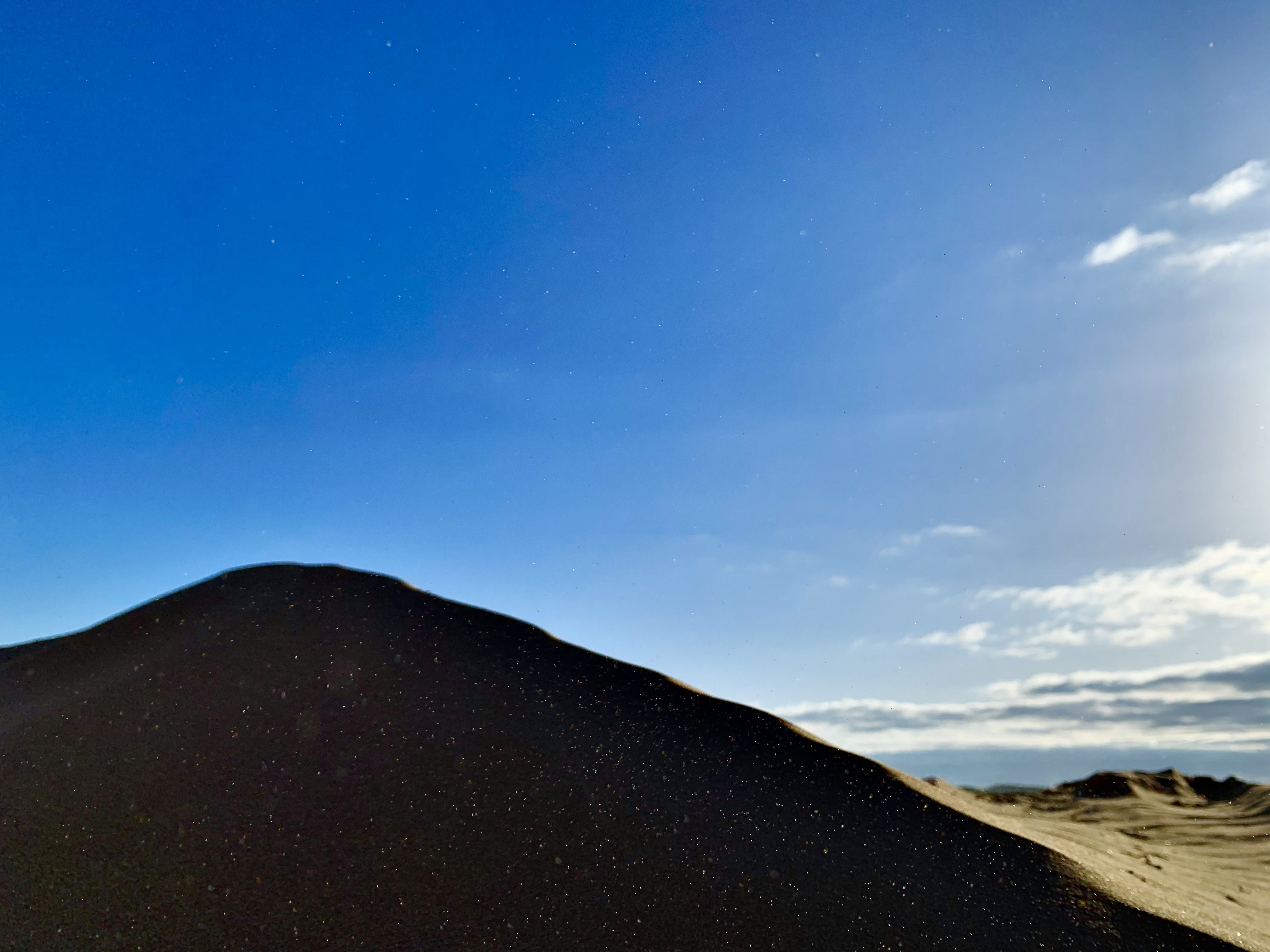 Dark sand dune contrasting against a bright blue sky and scattered clouds.
