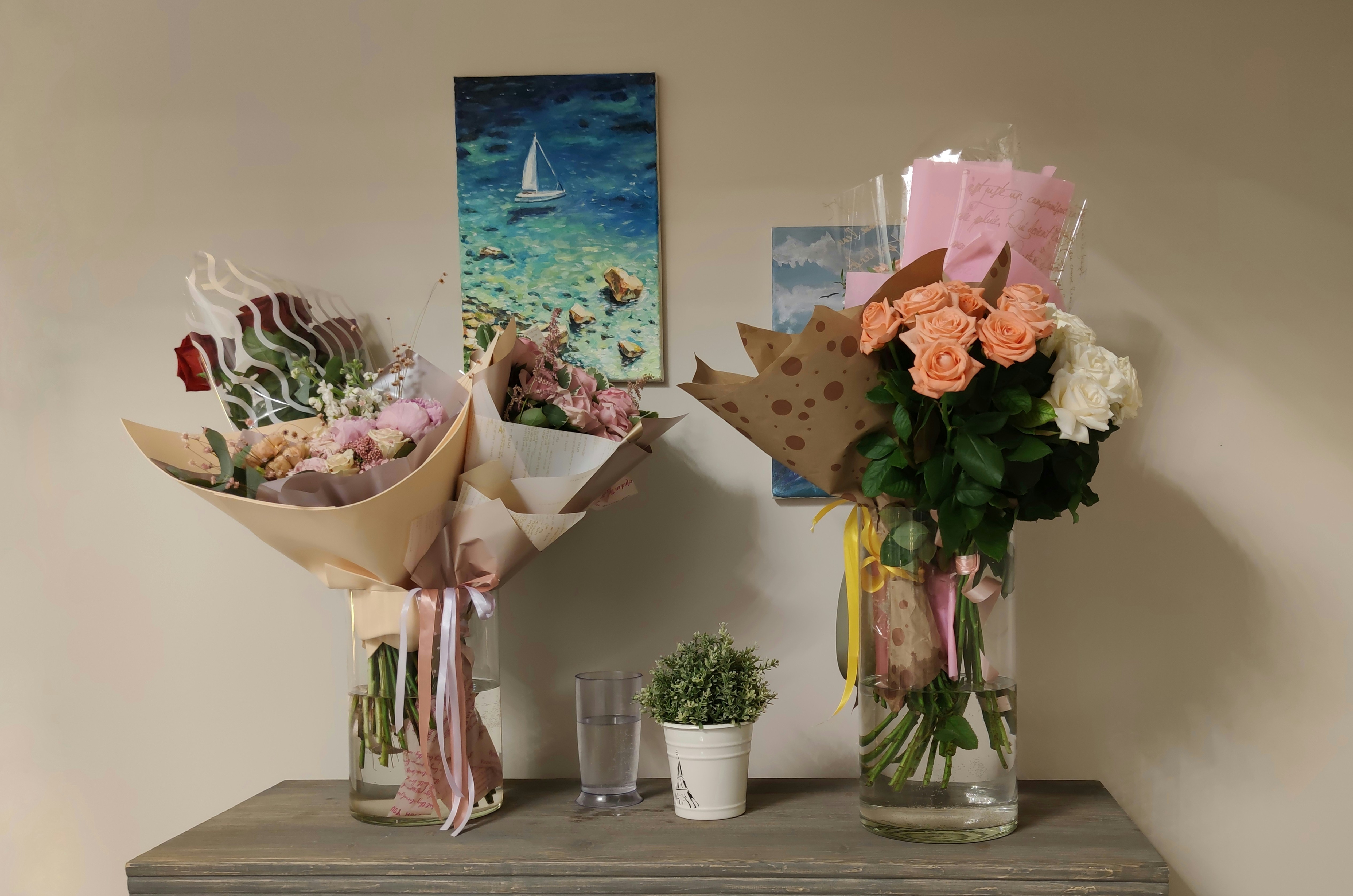 Bouquets in glass vases arranged on a wooden table with a small potted plant and water glass against a beige wall; a floral display with pastel wrapping and a backdrop featuring seaside artwork.