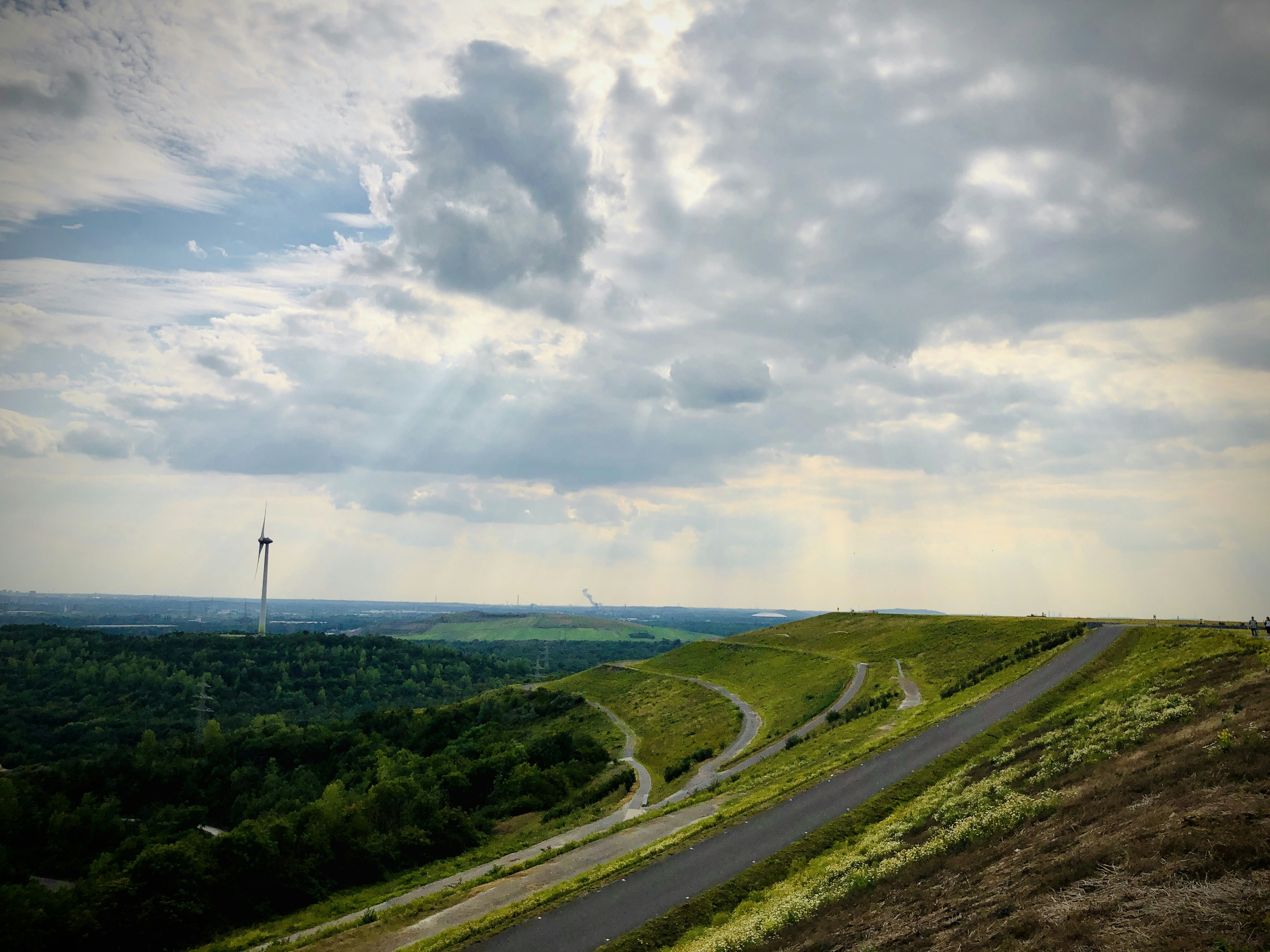 Curving road through lush green hills under a cloudy sky with scattered sunlight.