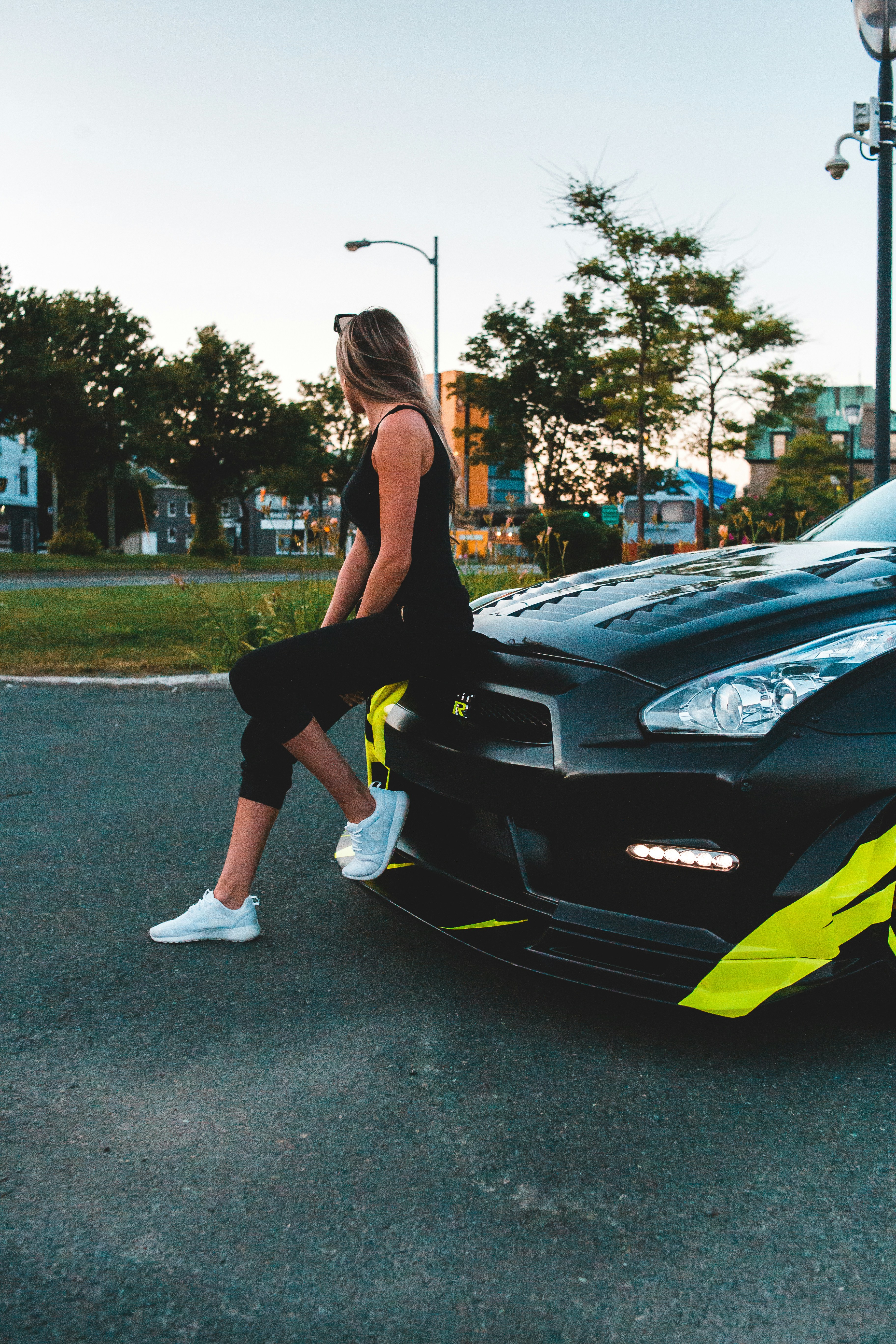 Neon-Tethered Pause: A woman in black perched on a neon-yellow sports sedan in a quiet parking lot