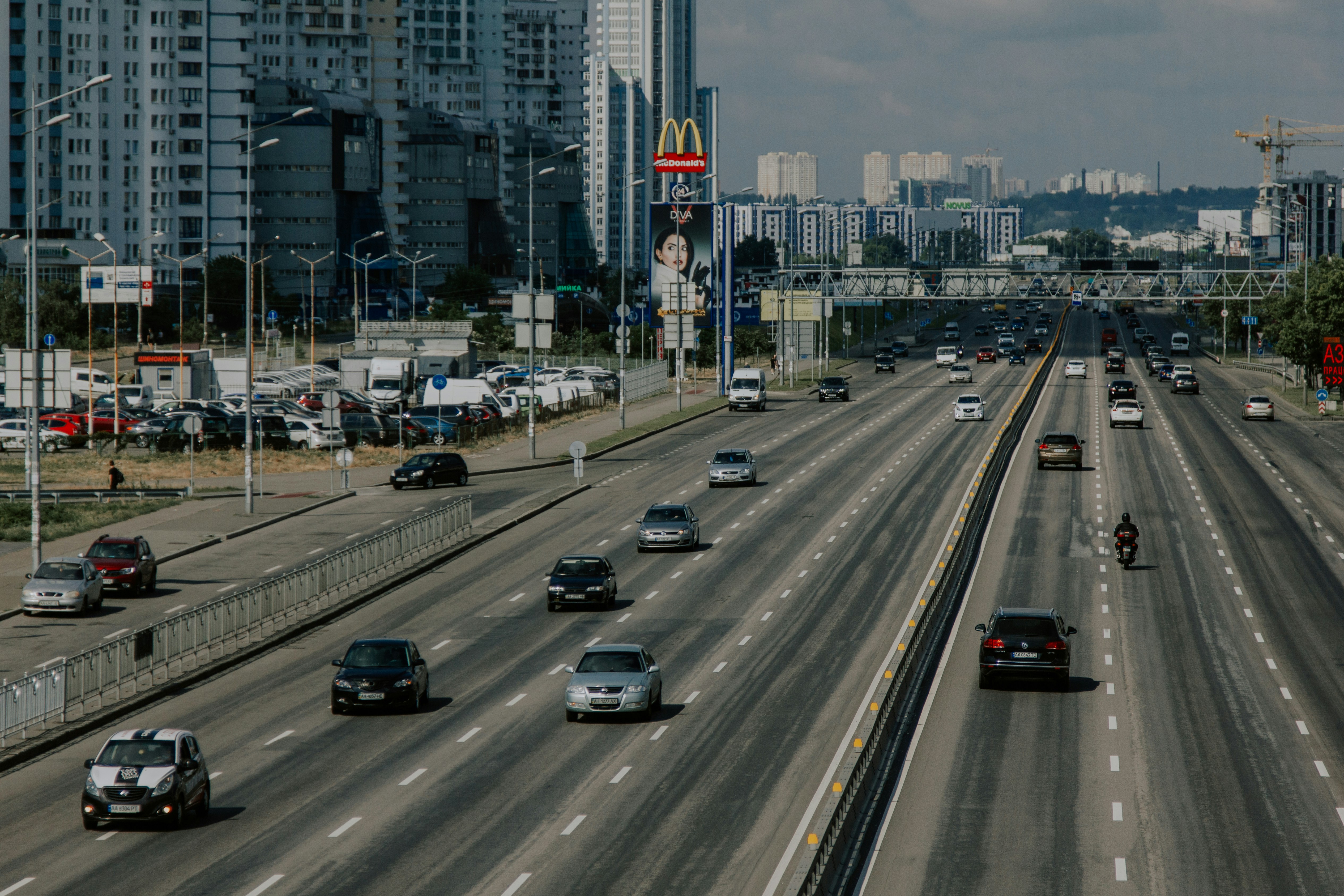 Wide highway with multiple lanes and cars, flanked by tall buildings under a cloudy sky.