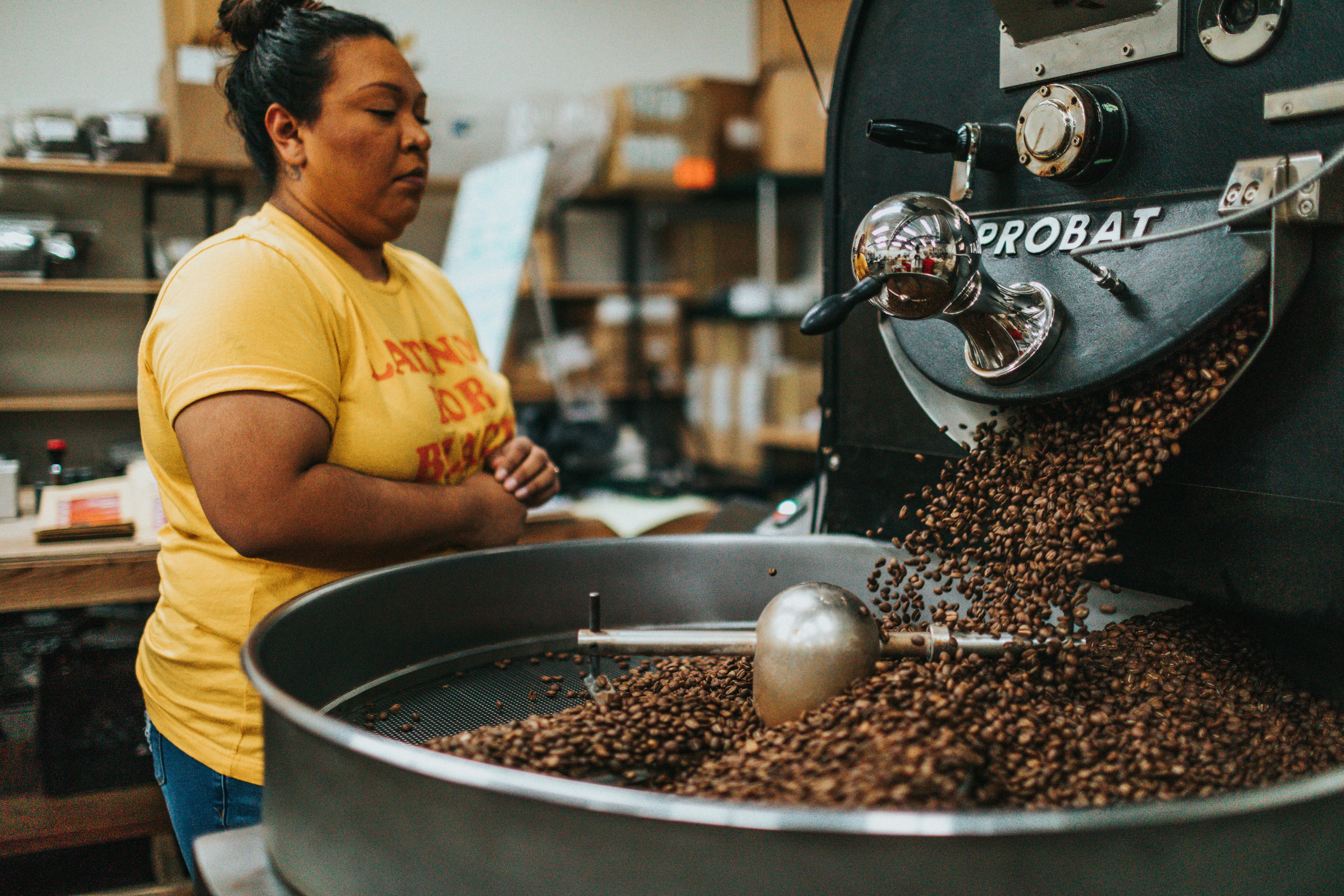 A woman skillfully operates a coffee roaster, with freshly roasted beans cascading into a large drum. The scene captures the essence of artisanal coffee production.