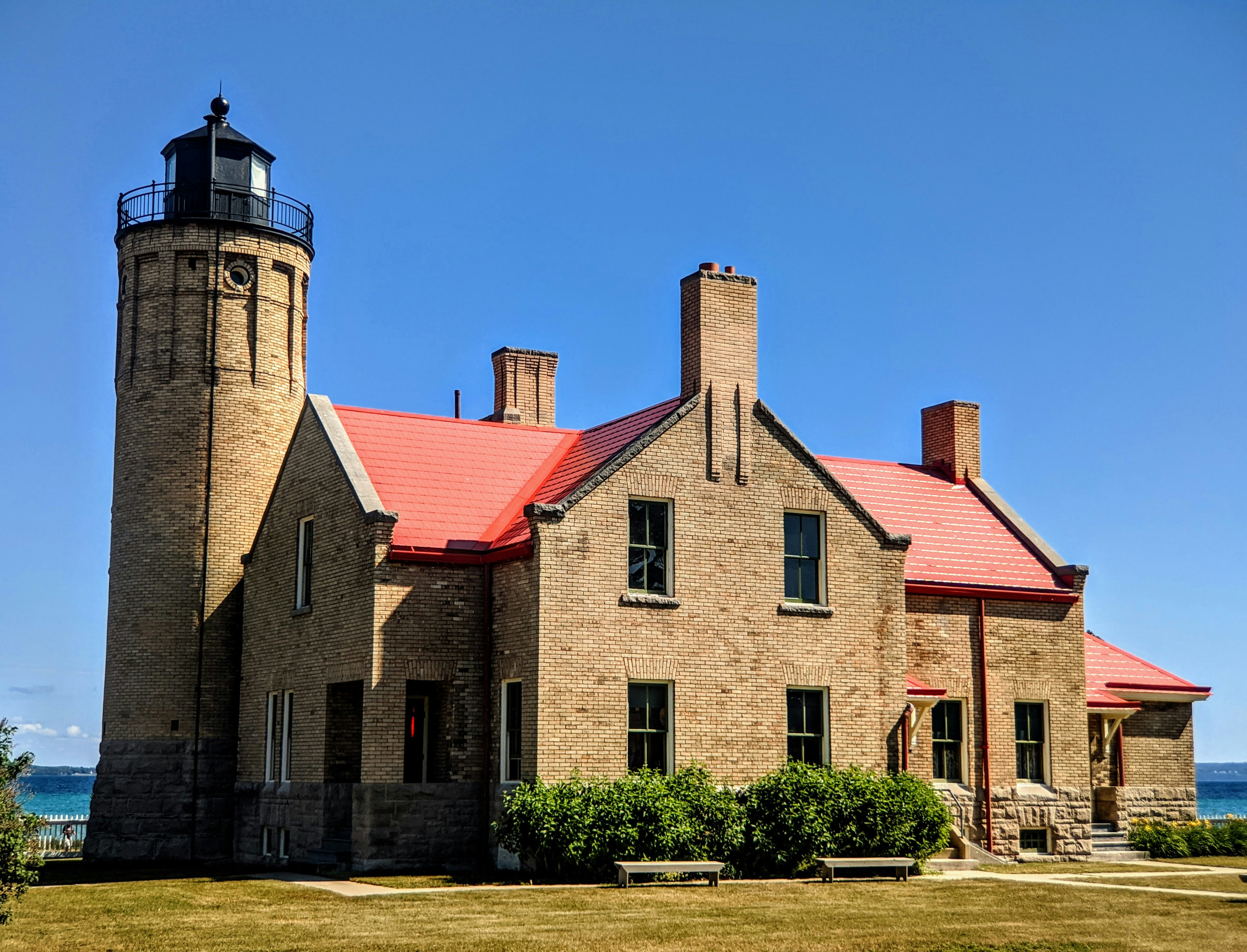 Historic lighthouse with a red roof and stone structure, surrounded by green landscaping and a clear blue sky.