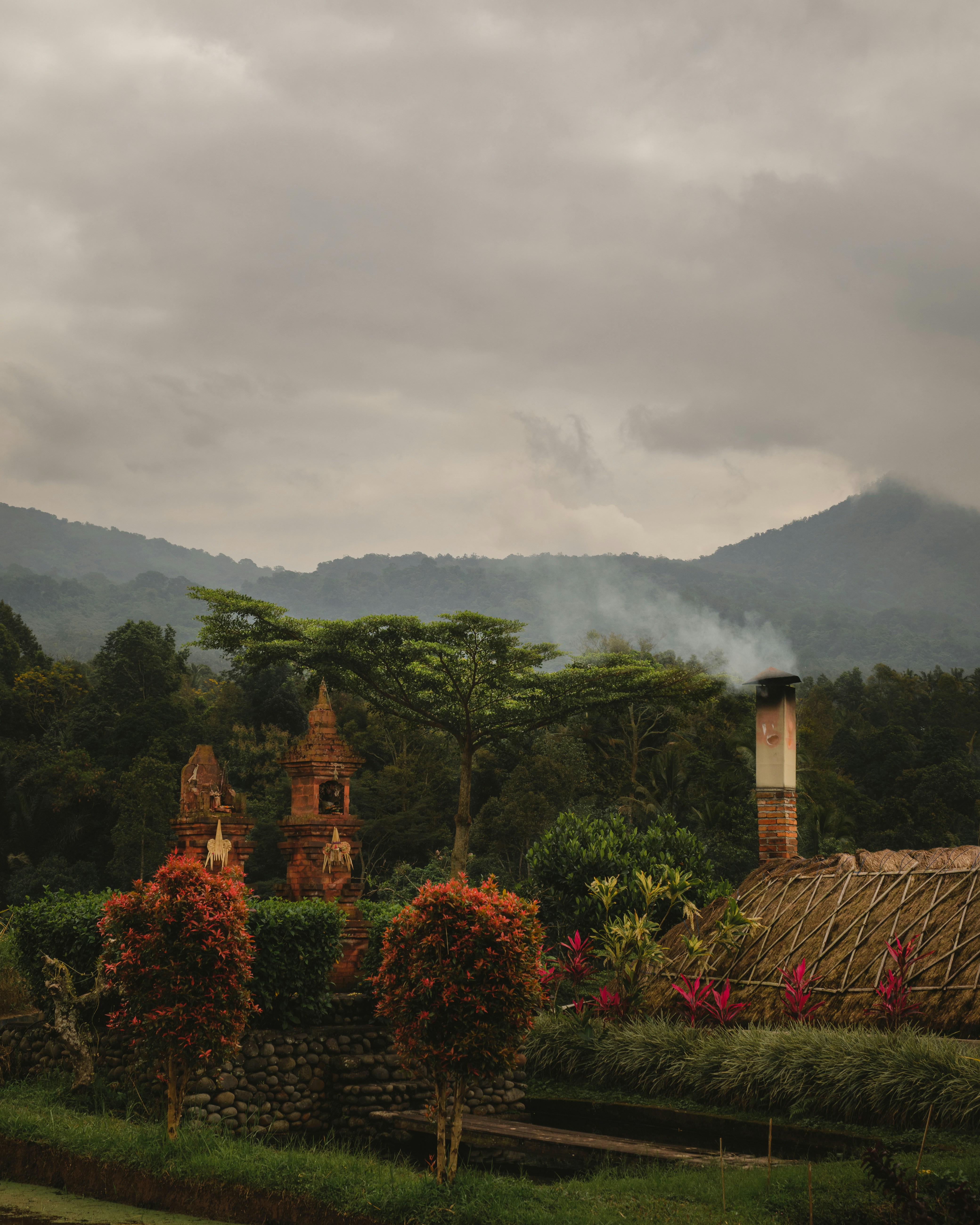 Ancient temple structures nestled among lush greenery, with smoke rising from a chimney against a backdrop of rolling hills and overcast skies.