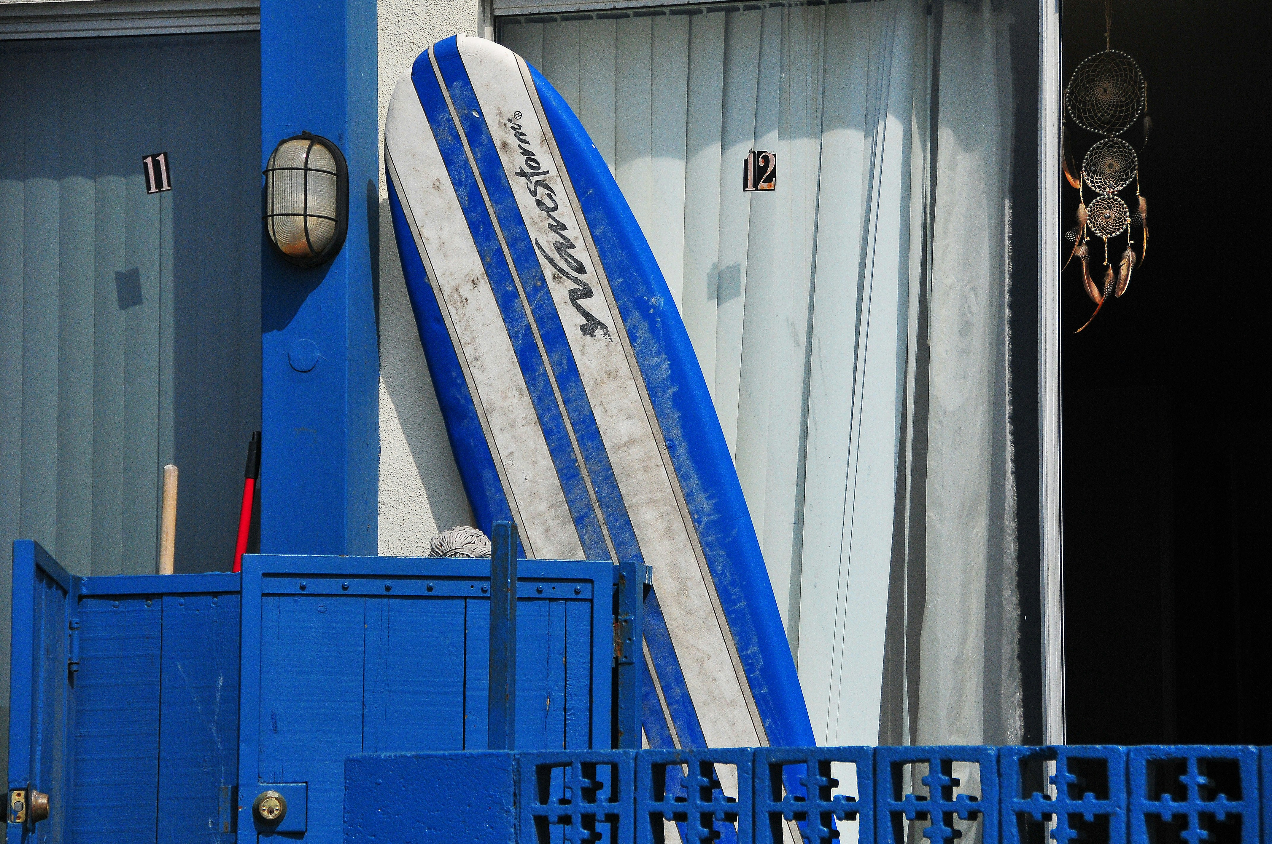 A blue surfboard rests against a vibrant blue wall, hinting at a laid-back coastal lifestyle. Dreamcatchers hang nearby, adding a touch of artistic charm.