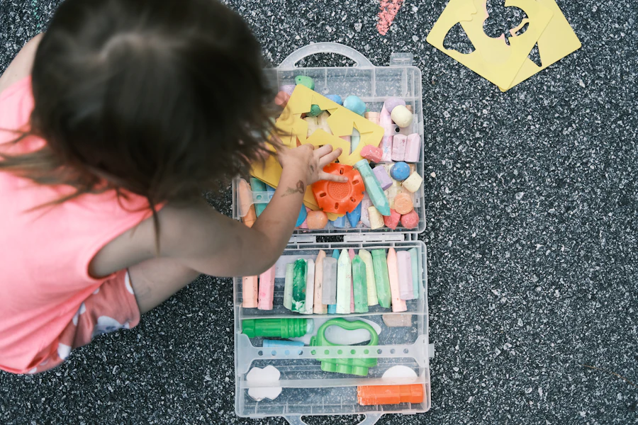 A child explores a transparent box filled with colorful chalk and stencils on a textured surface. The vibrant assortment invites imaginative play.